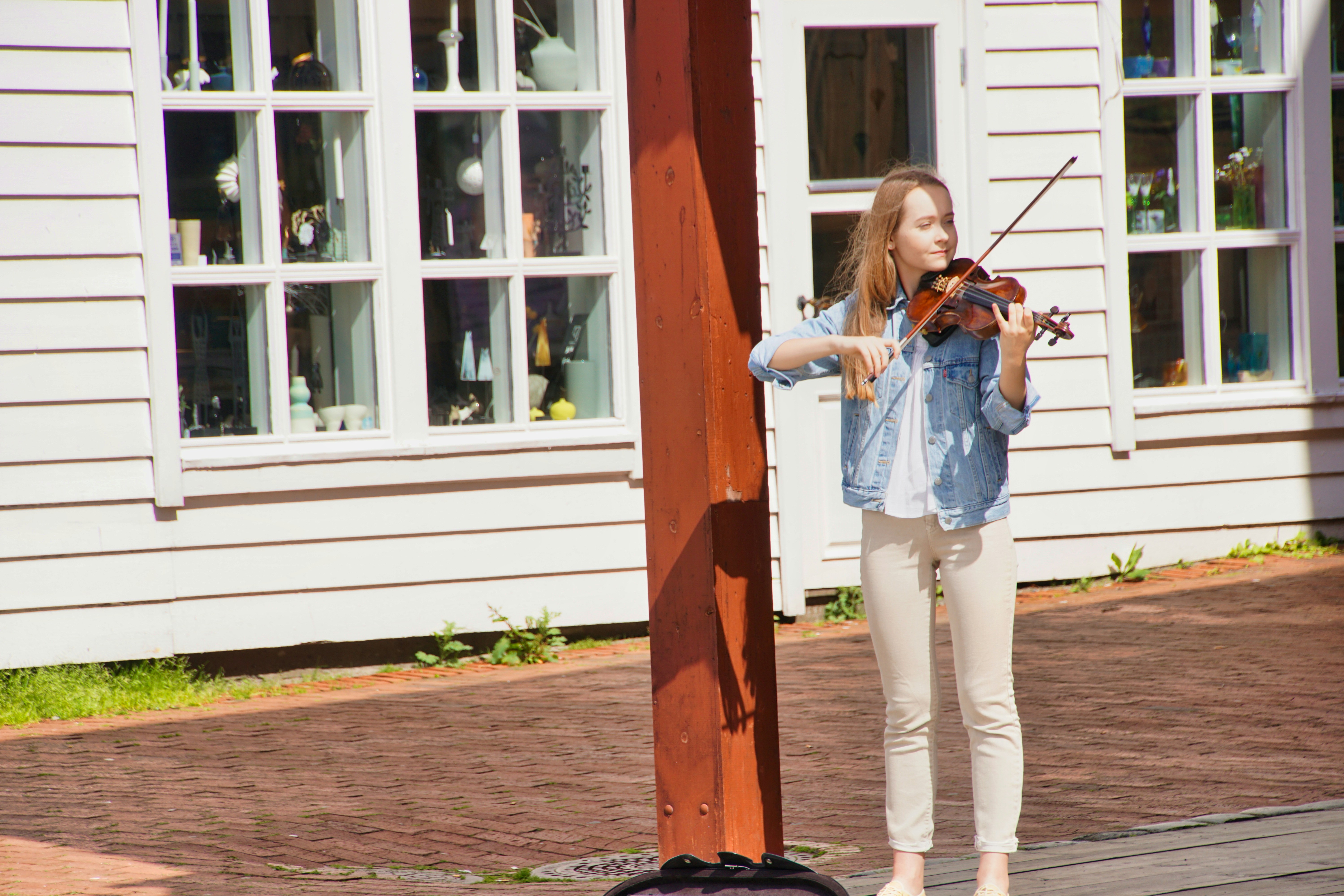 Young musician playing the violin outdoors, surrounded by quaint shopfronts. The scene captures a lively moment of artistic expression.
