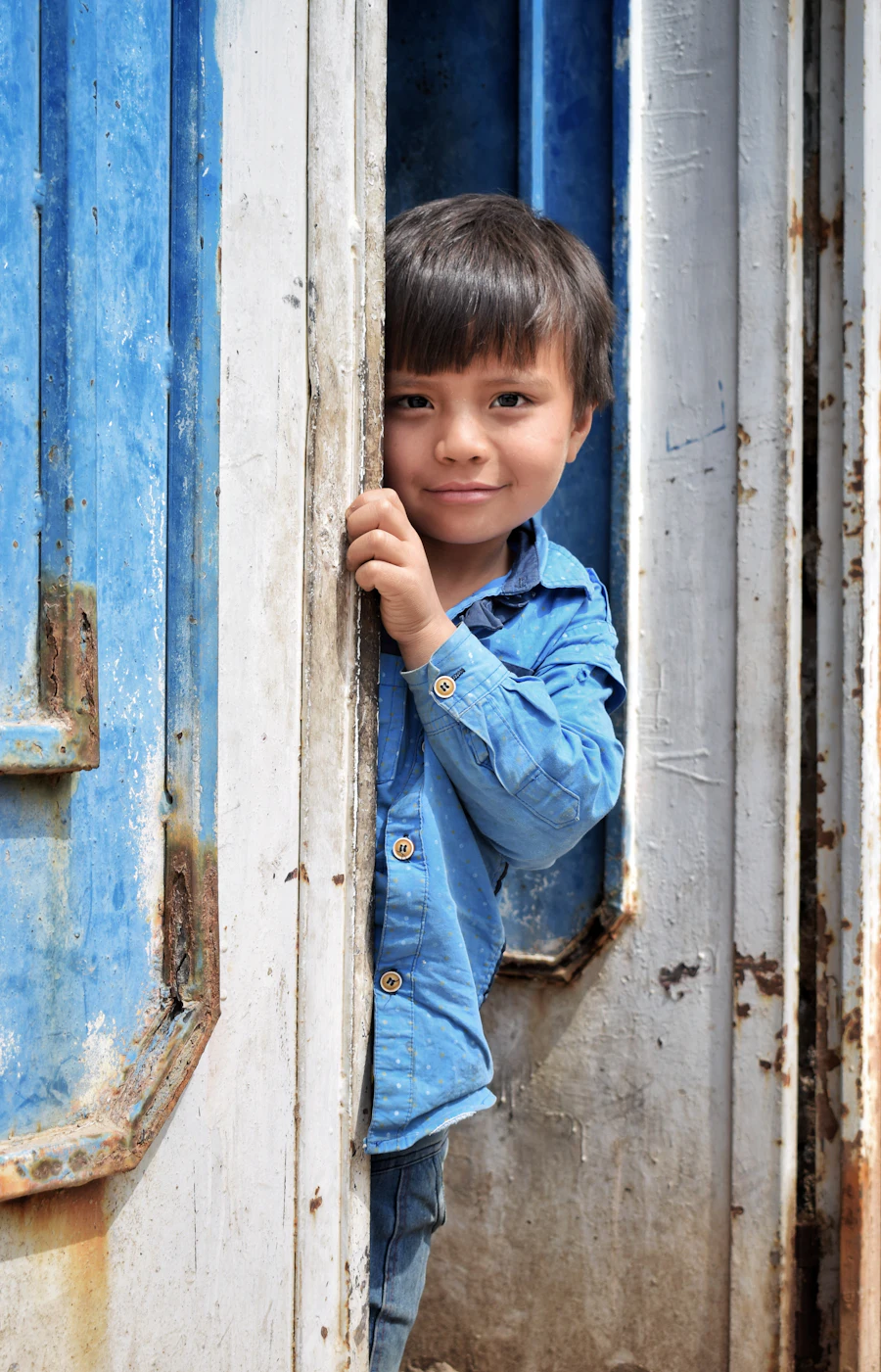 Young boy in a blue shirt peeks through a narrow gap in peeling blue and white metal doors, holding the edge with one hand. Rust and chipped paint create a worn, textured backdrop.
