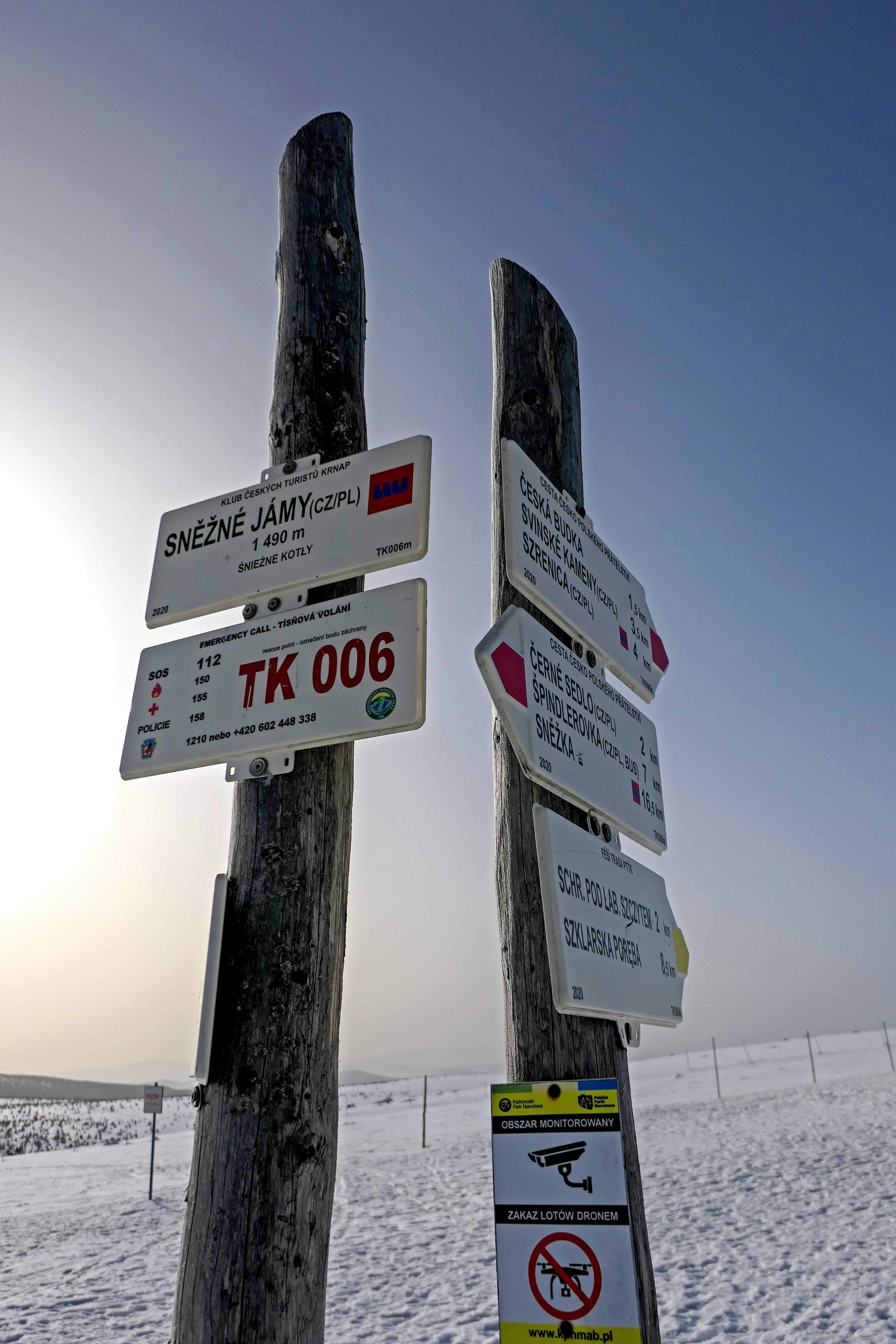 Two weathered wooden signposts displaying trail information and directions in a snowy landscape. The sun casts a soft glow in the background.