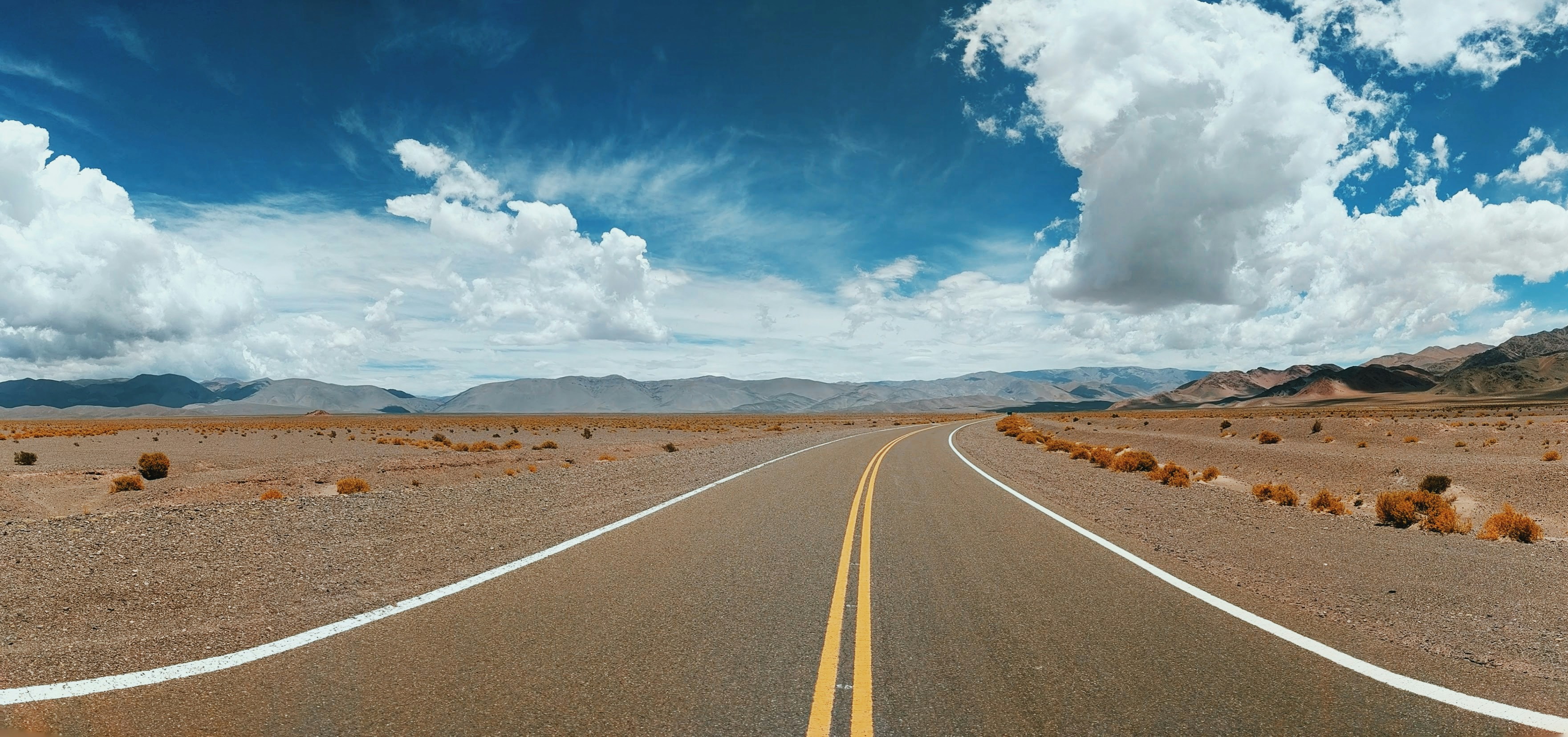 Lonely asphalt road stretching through arid desert landscape under expansive blue sky with scattered clouds.