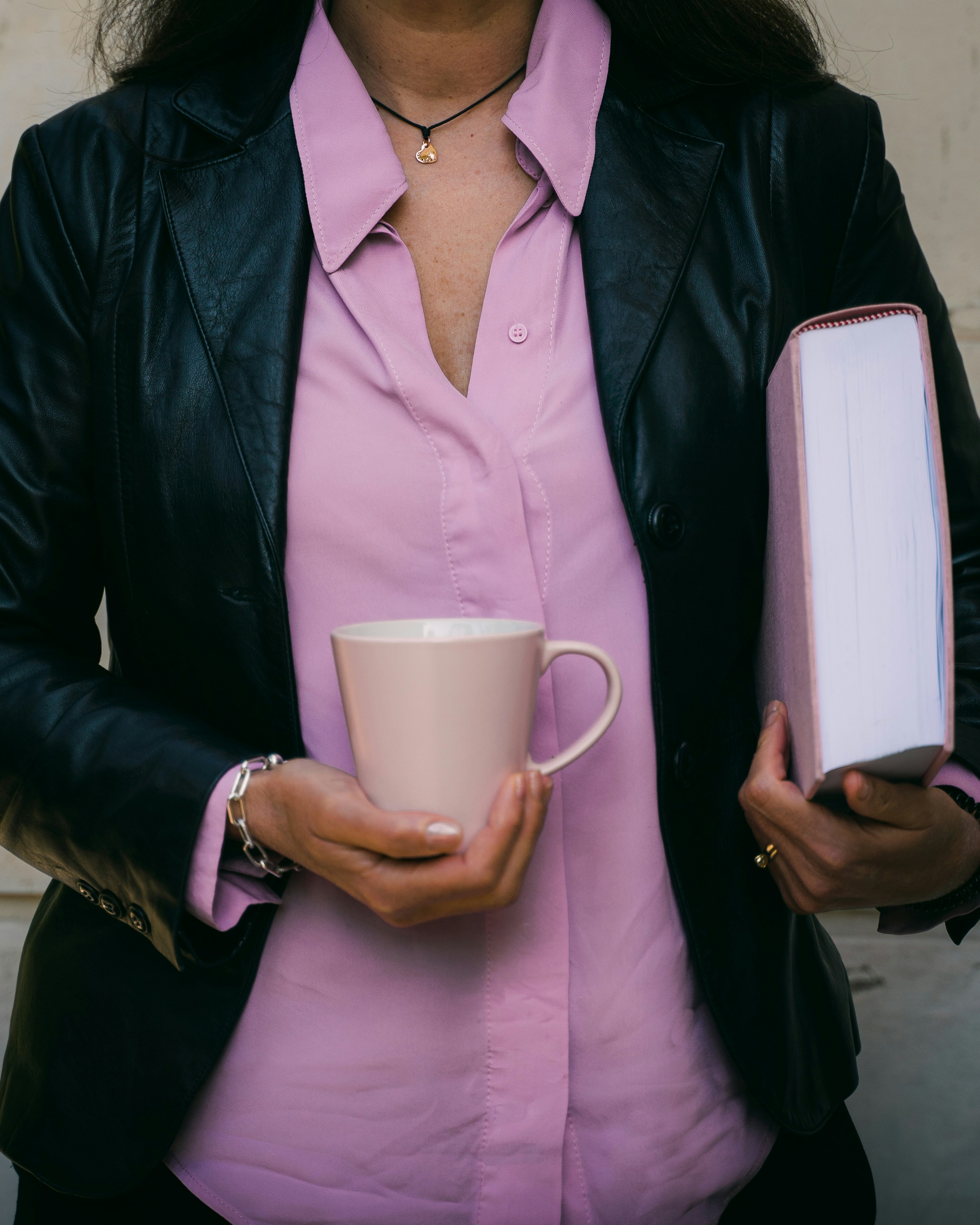 A person in a black leather jacket holds a pink mug and a book, embodying a blend of sophistication and casual comfort.