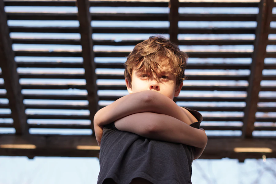 A young boy stands with arms crossed, gazing thoughtfully, framed by a pattern of shadows from overhead slats.