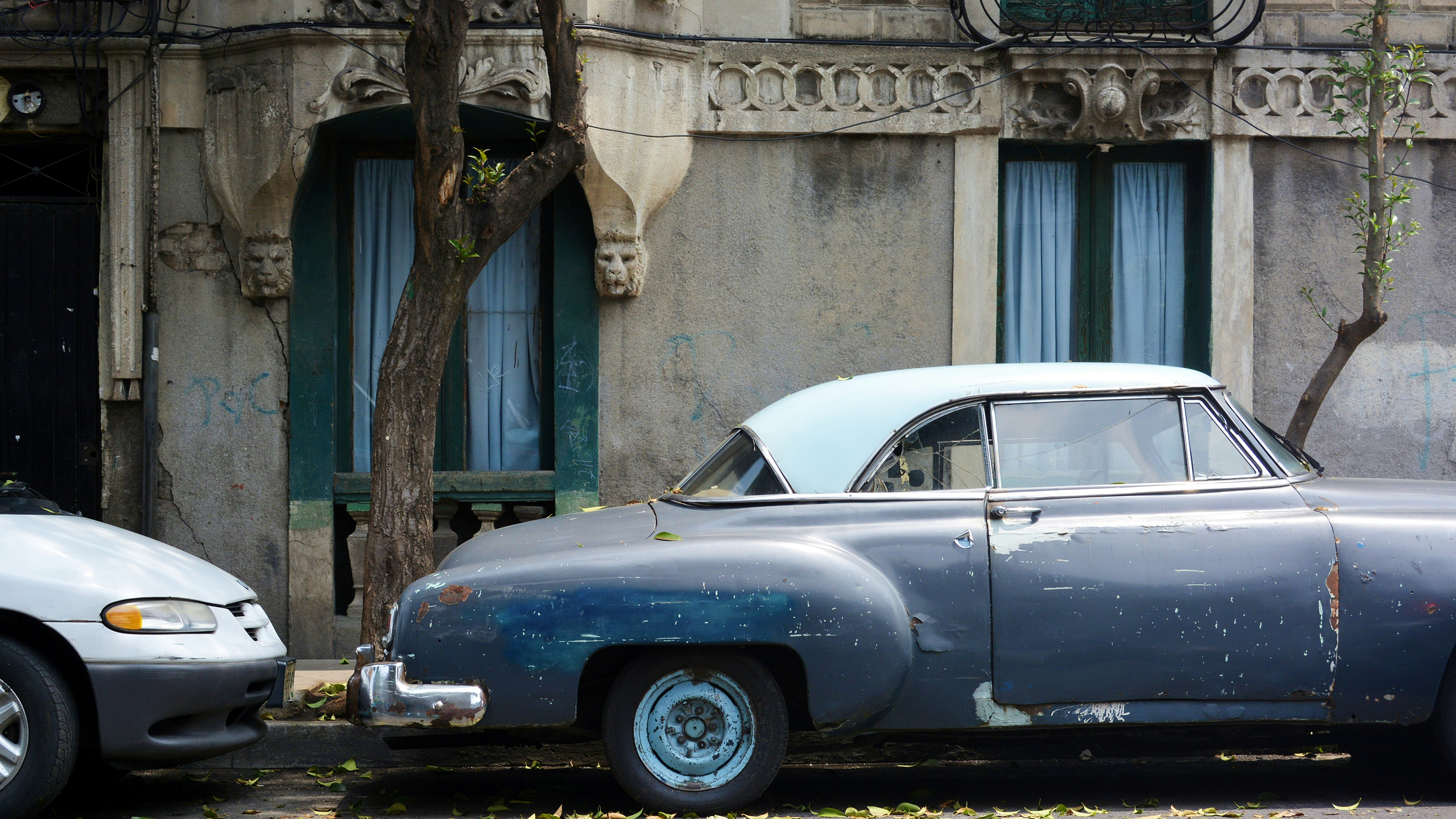 A vintage car, weathered and charming, parked beside a modern vehicle in front of an old building with decorative architecture.