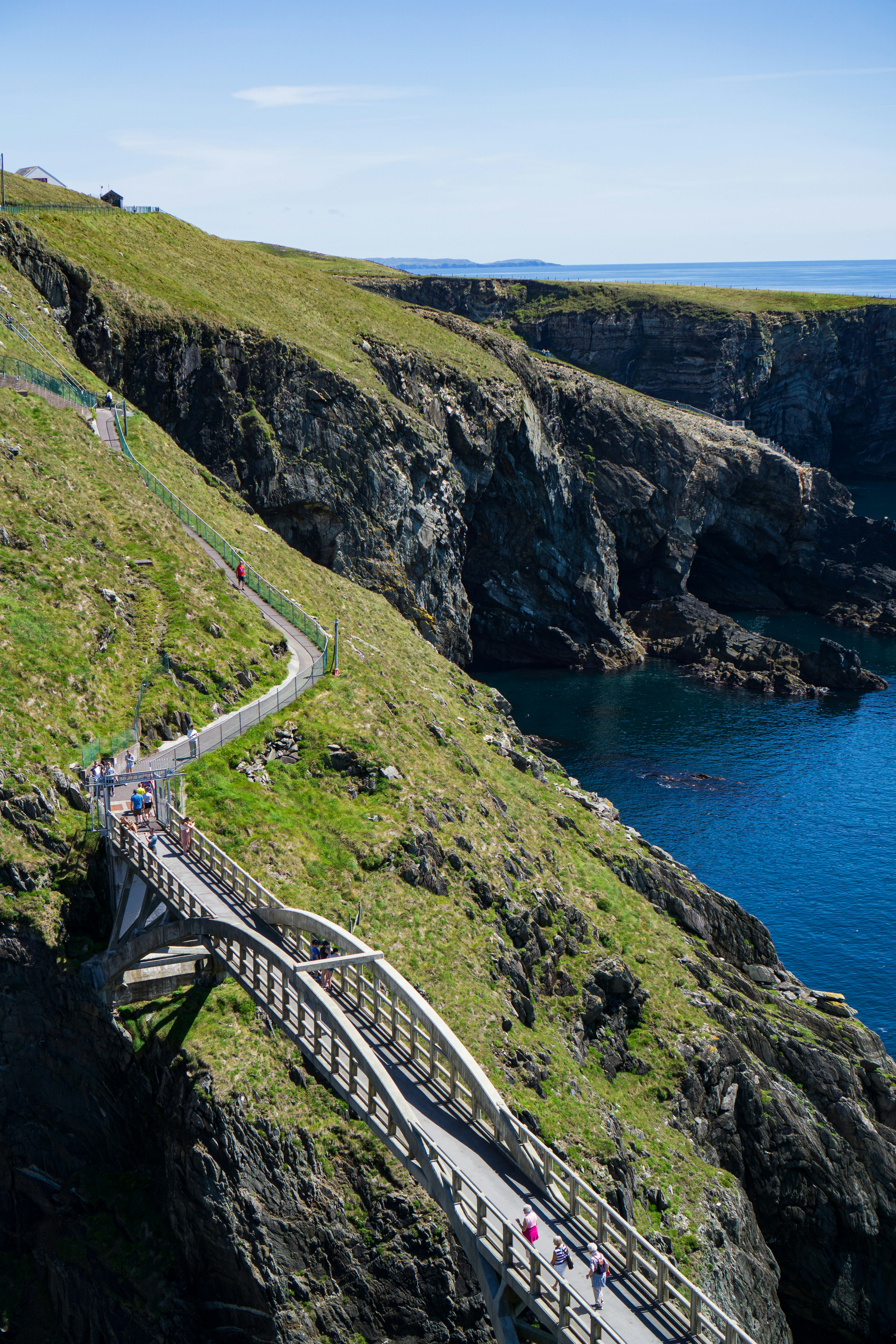 A wooden walkway meanders along steep cliffs, leading to the vibrant blue waters below, surrounded by lush greenery.