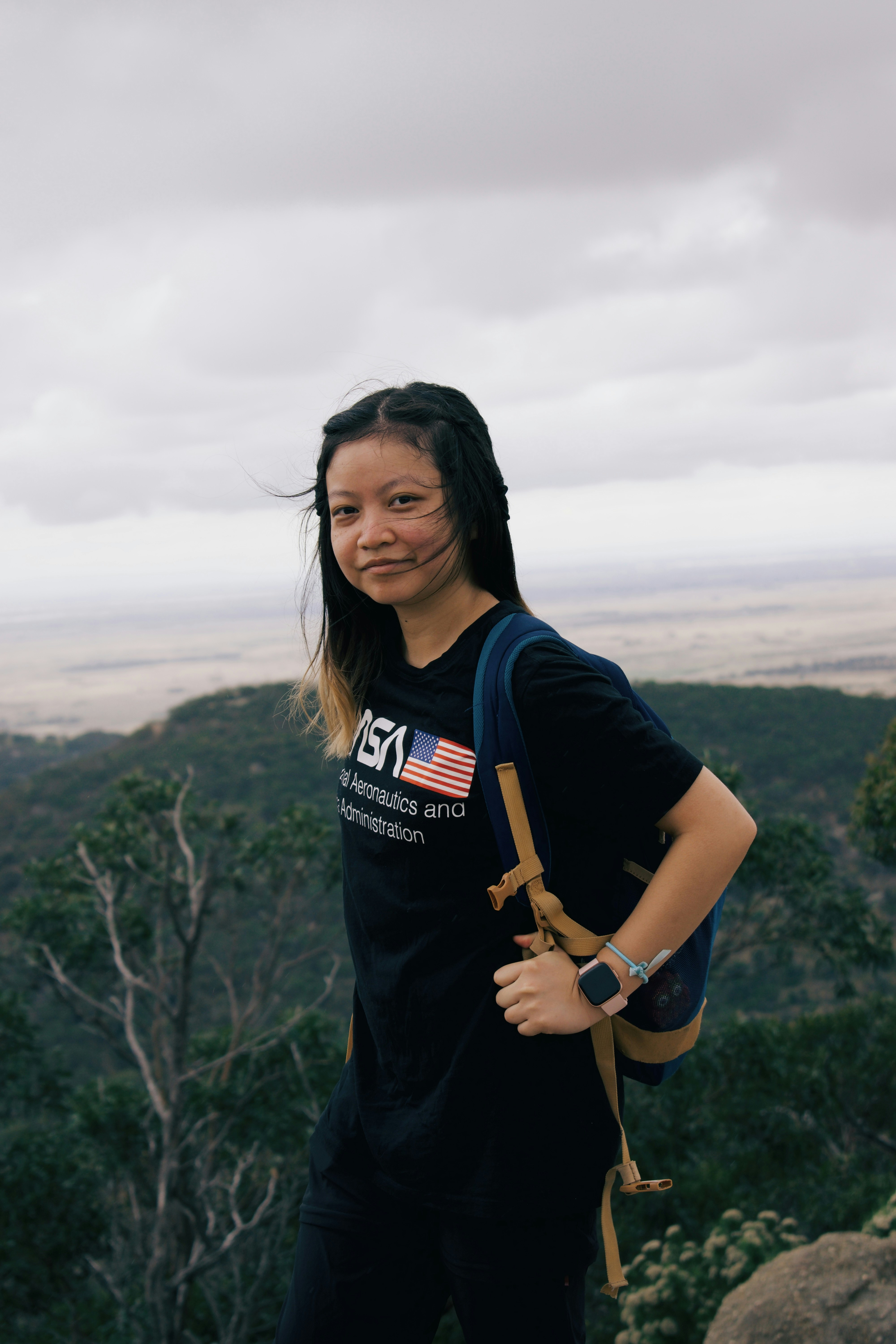 A young woman stands confidently on a mountain overlook, dressed in a NASA-themed shirt, with a backpack and a scenic backdrop of rolling hills and cloudy skies.