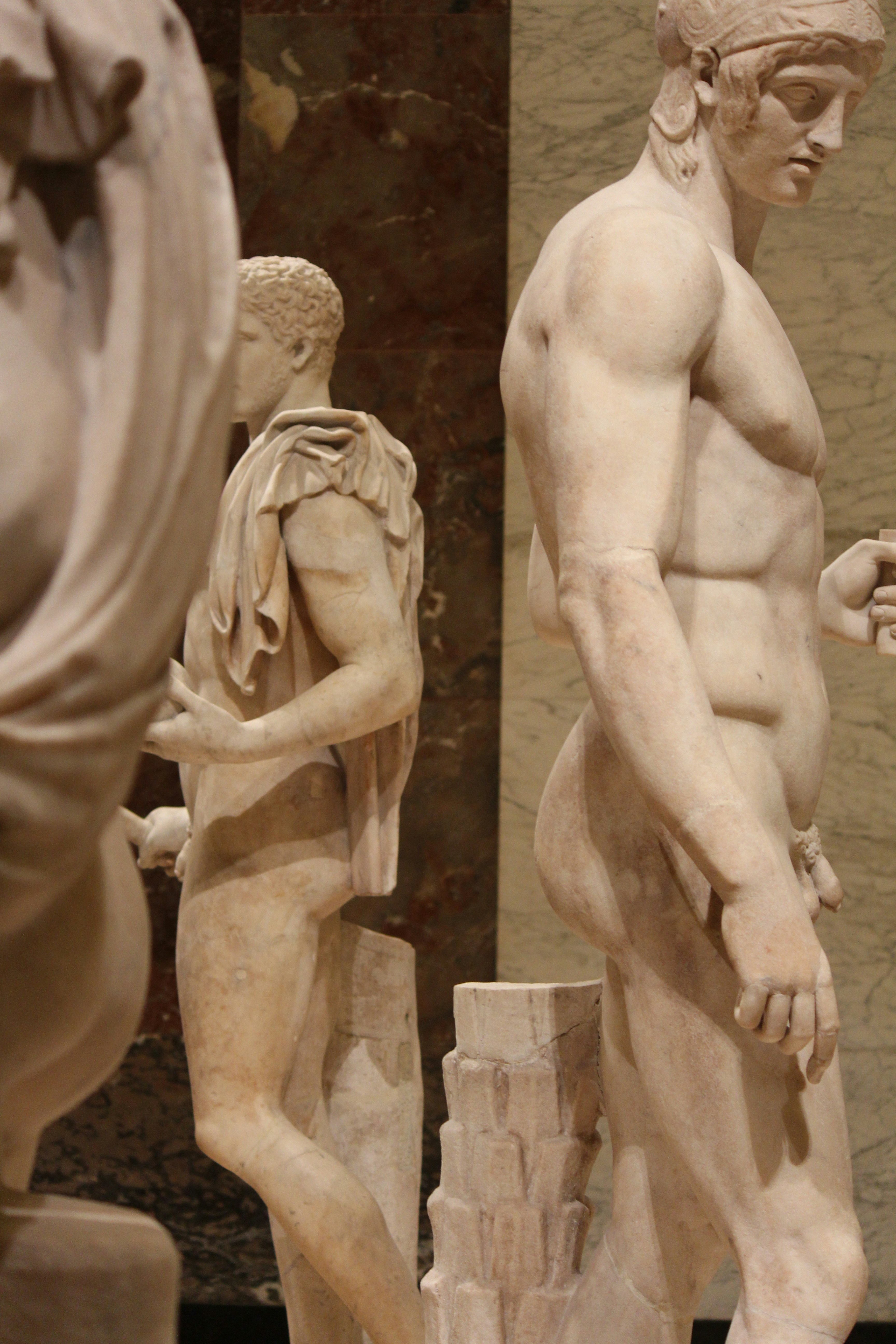 Two classical marble statues stand in a museum setting, showcasing intricate details and historical artistry. The focus is on their graceful forms and expressions.