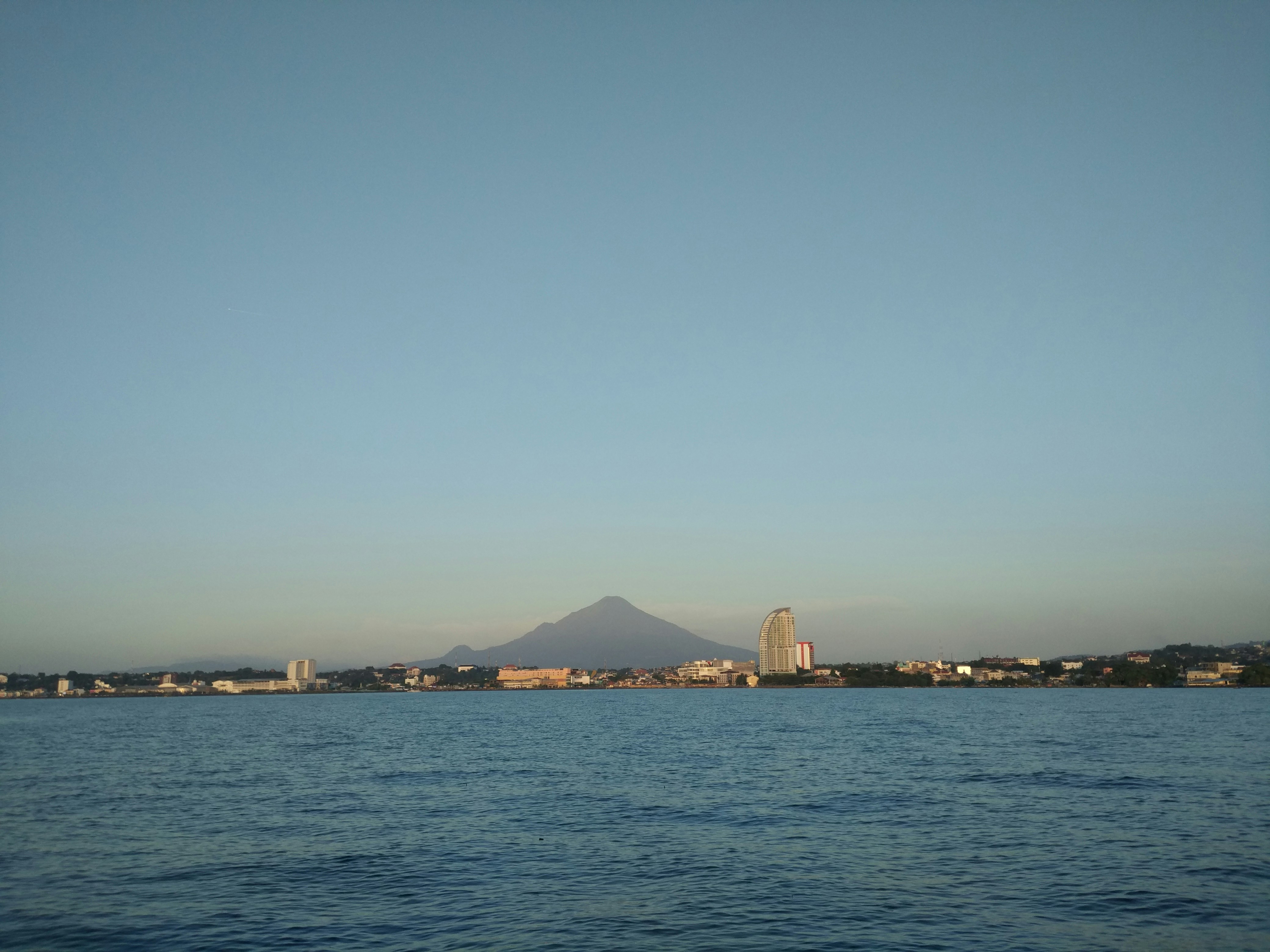 Majestic volcano looms over a tranquil waterfront cityscape at dusk, with calm waters reflecting the soft hues of the sky.