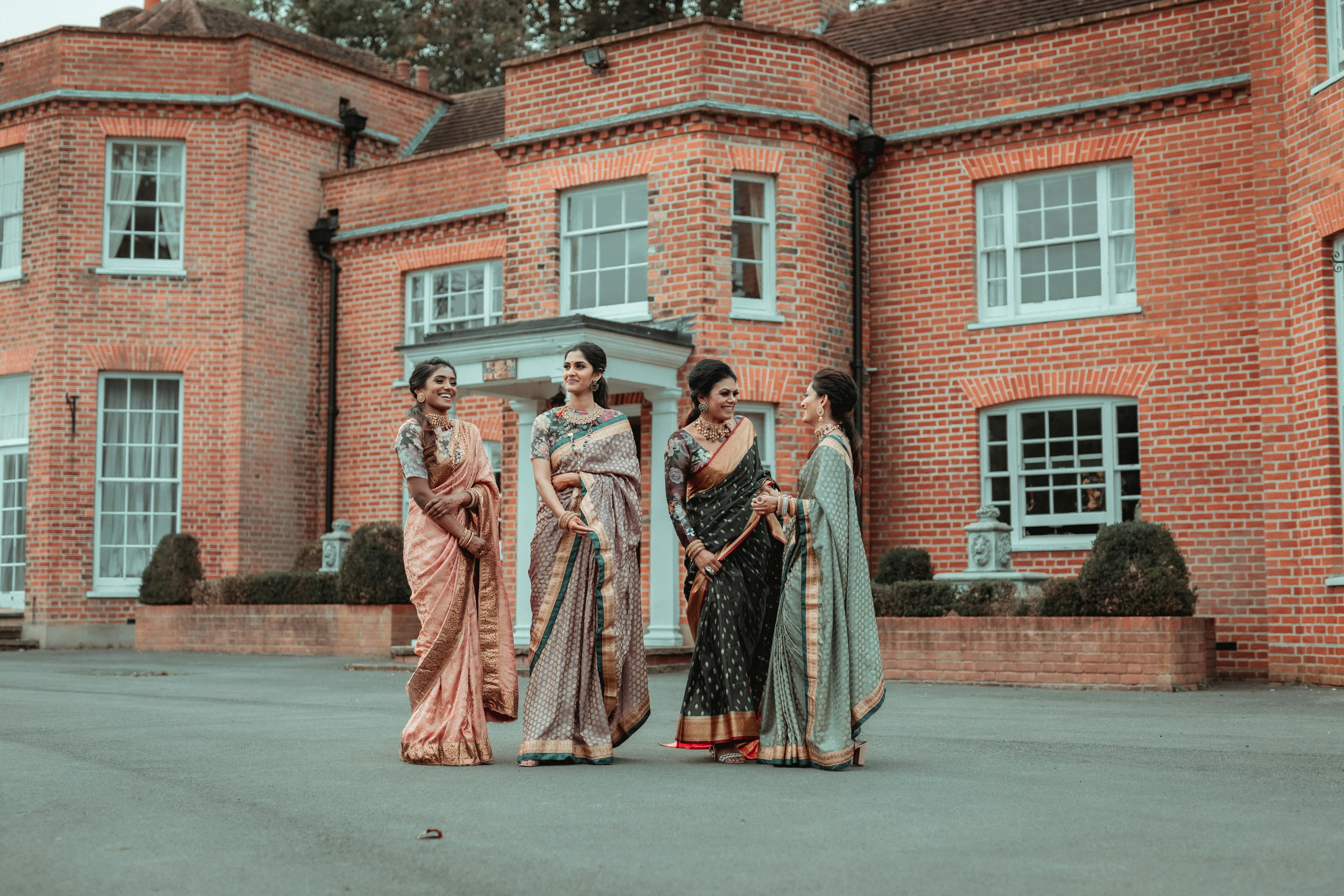 Four women in vibrant saris stand in front of a grand brick building, engaged in conversation.