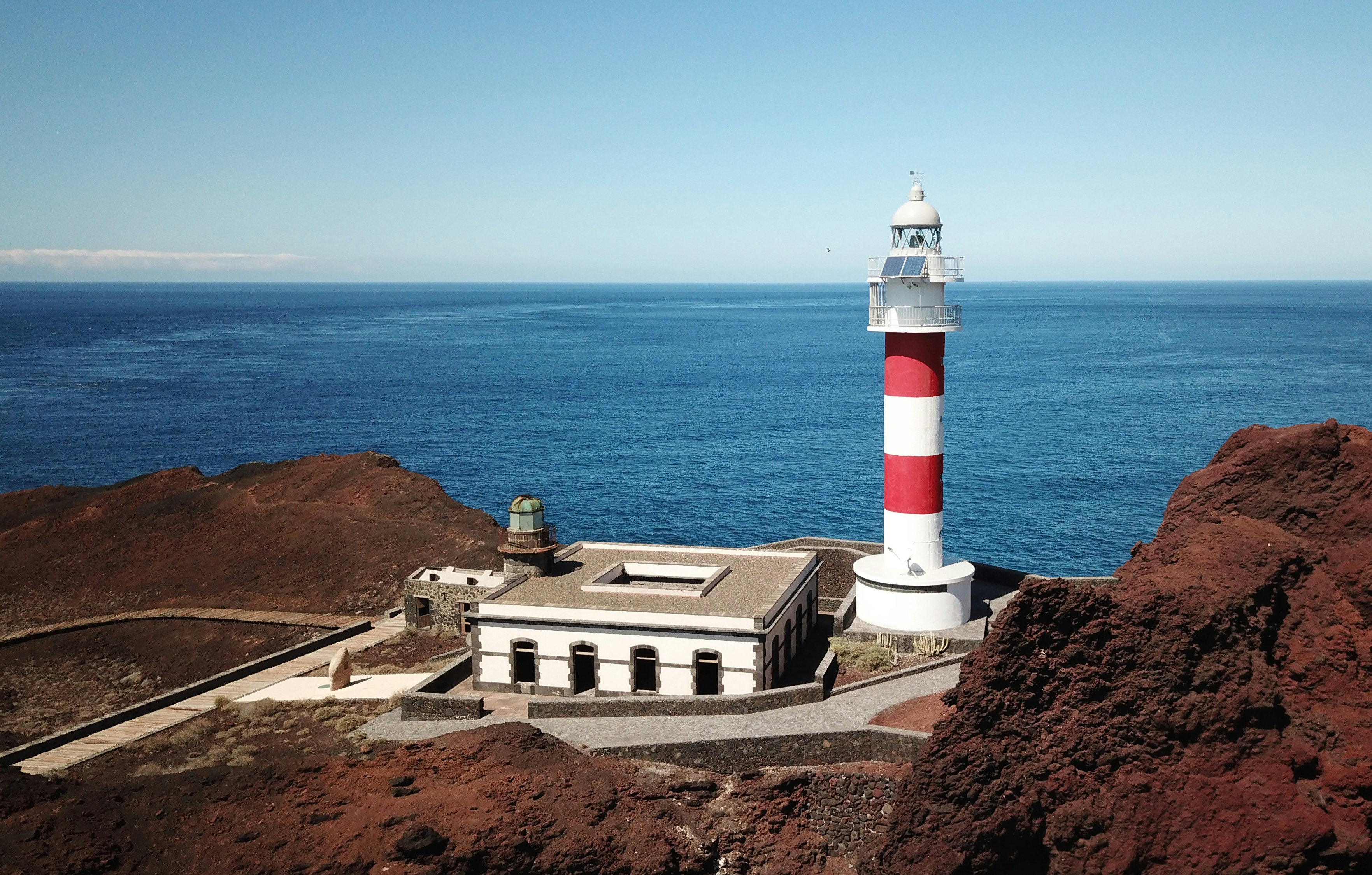A striking lighthouse stands on rugged terrain, overlooking the vast ocean under a clear blue sky. The vibrant red and white stripes contrast beautifully with the surrounding landscape.
