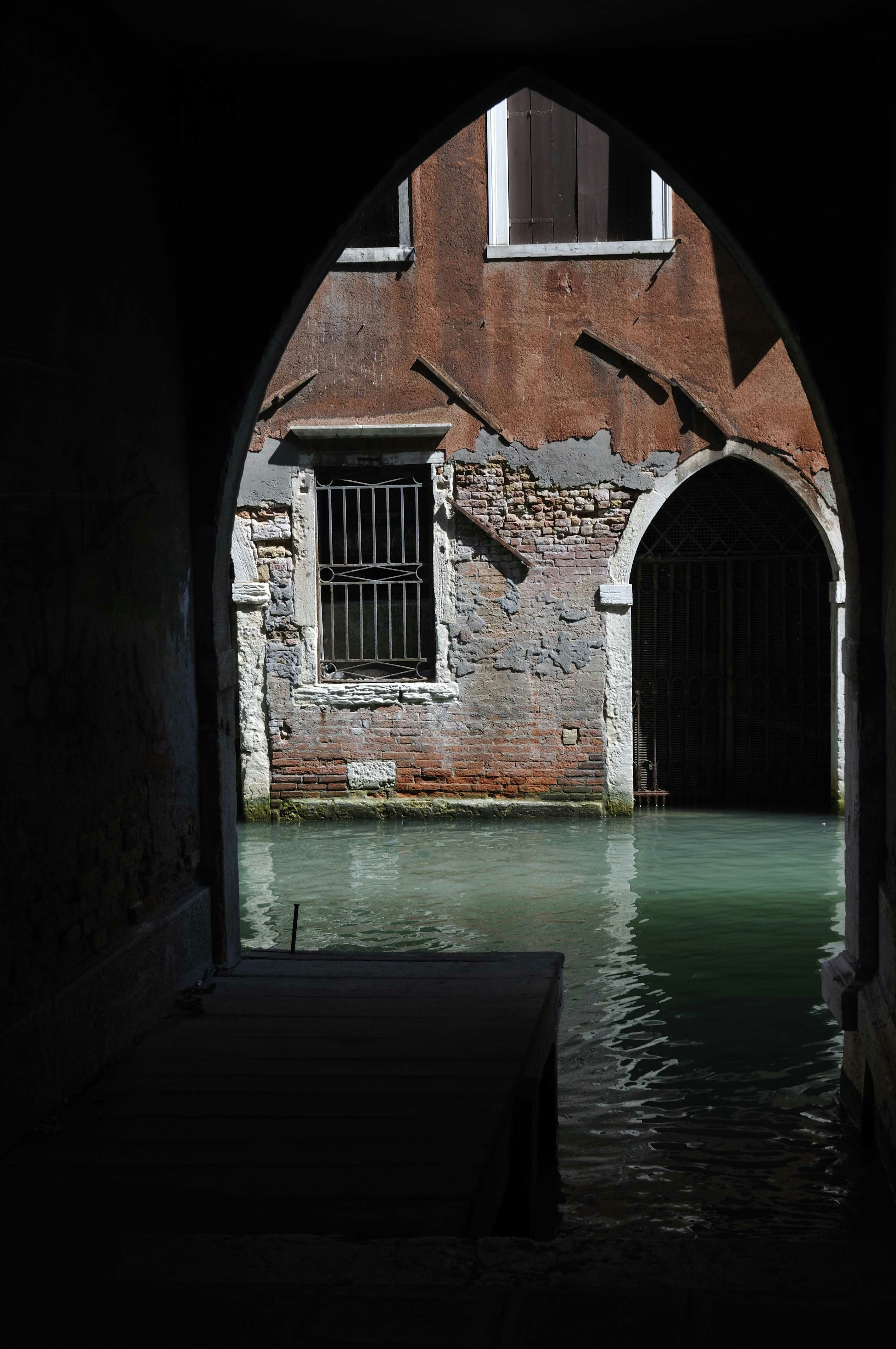 Weathered brick walls frame a serene waterway, revealing a glimpse of Venetian architecture through a darkened archway.