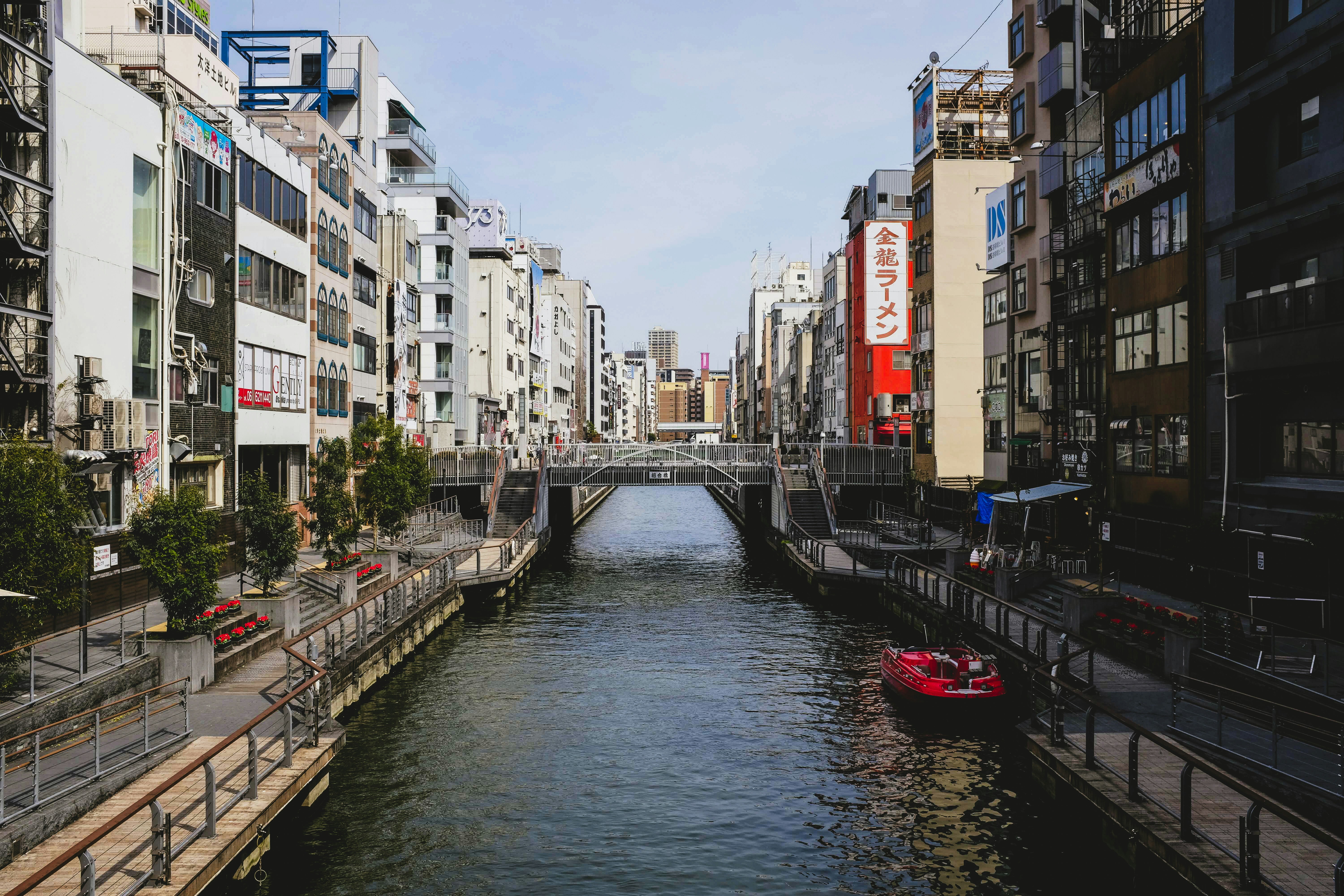 A tranquil canal flows through a bustling cityscape, flanked by modern buildings and greenery. A small red boat adds a touch of color to the serene waters.