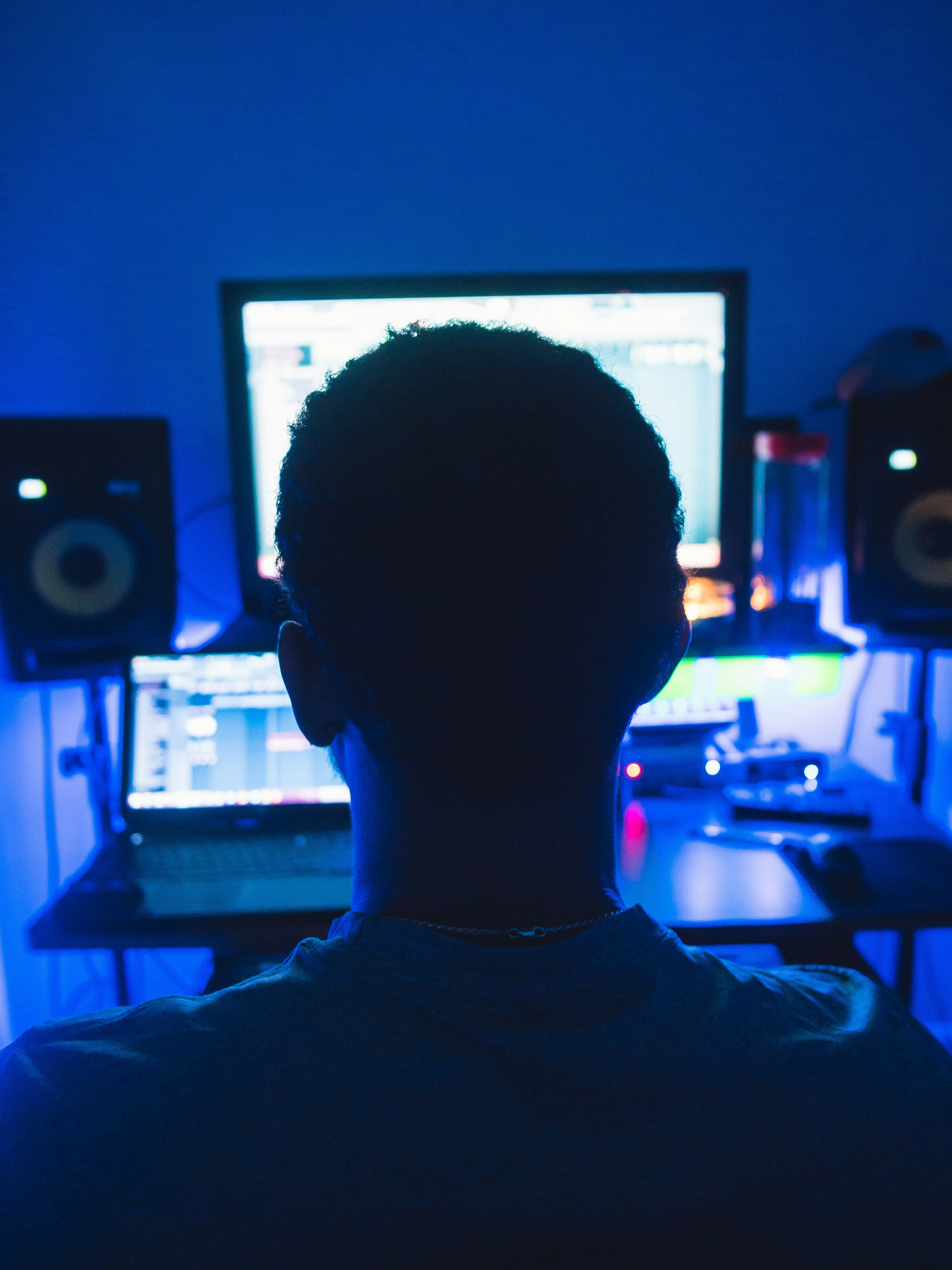 Silhouette of a person immersed in music production, surrounded by glowing monitors and speakers in a dimly lit studio.