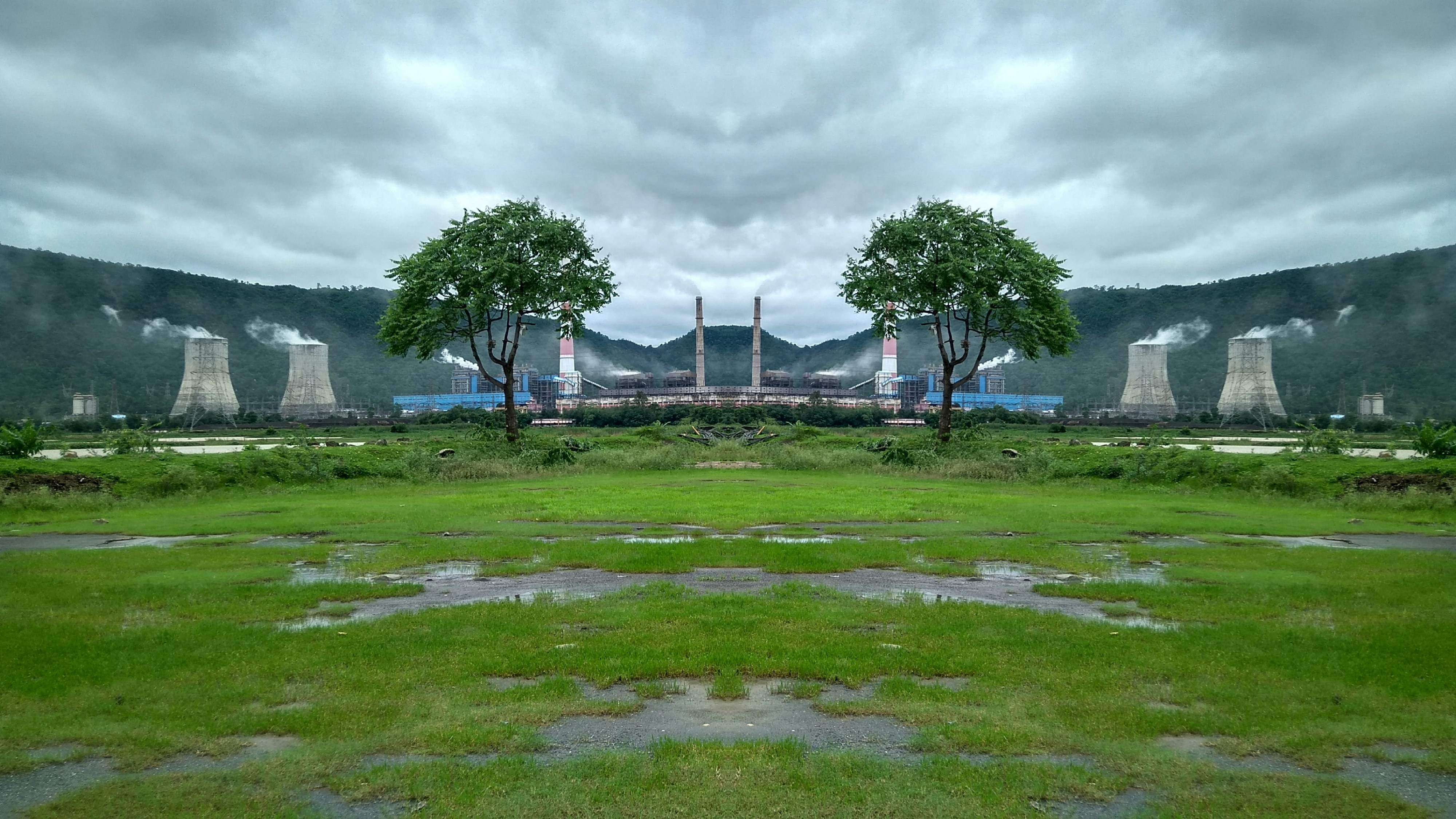 Landscape photograph showing two foreground trees framing a distant industrial complex with cooling towers under a cloudy sky.