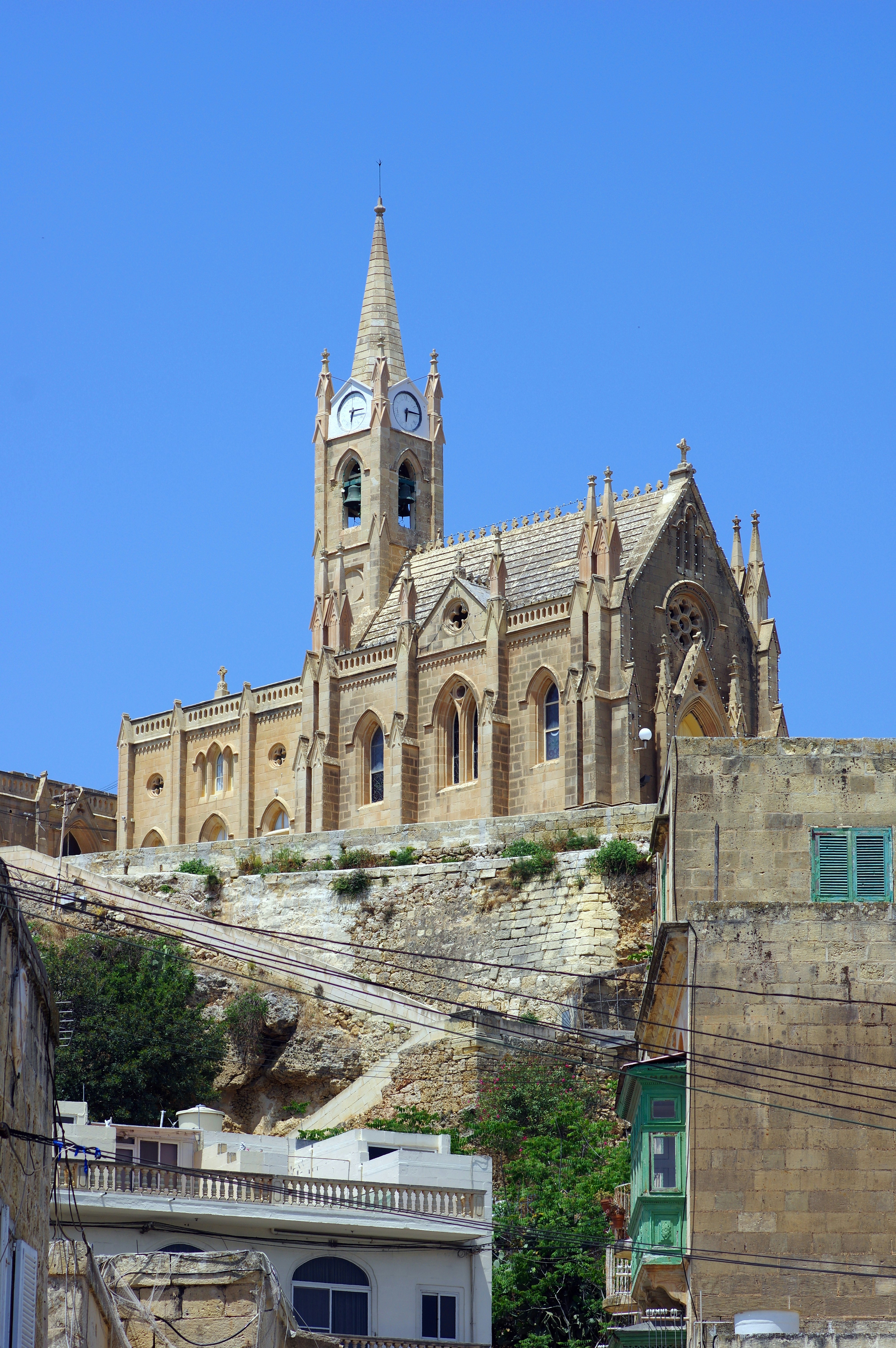 Stone Gothic church rises on a hillside above foreground buildings, with utility lines crossing the scene beneath a vivid blue sky.