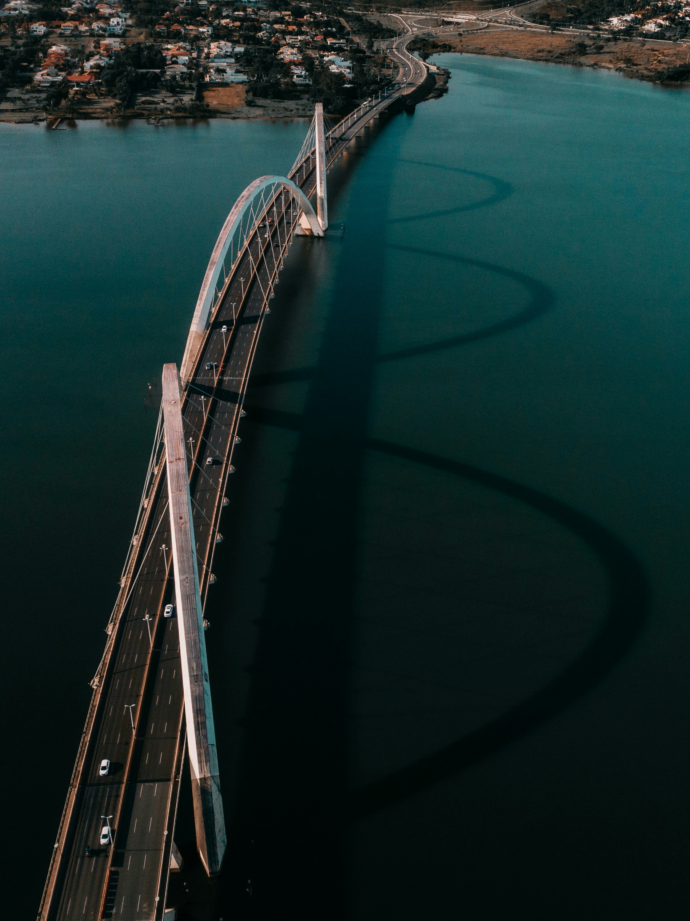 Aerial view of a modern bridge casting a long shadow over tranquil waters, with vehicles traversing its curved path.