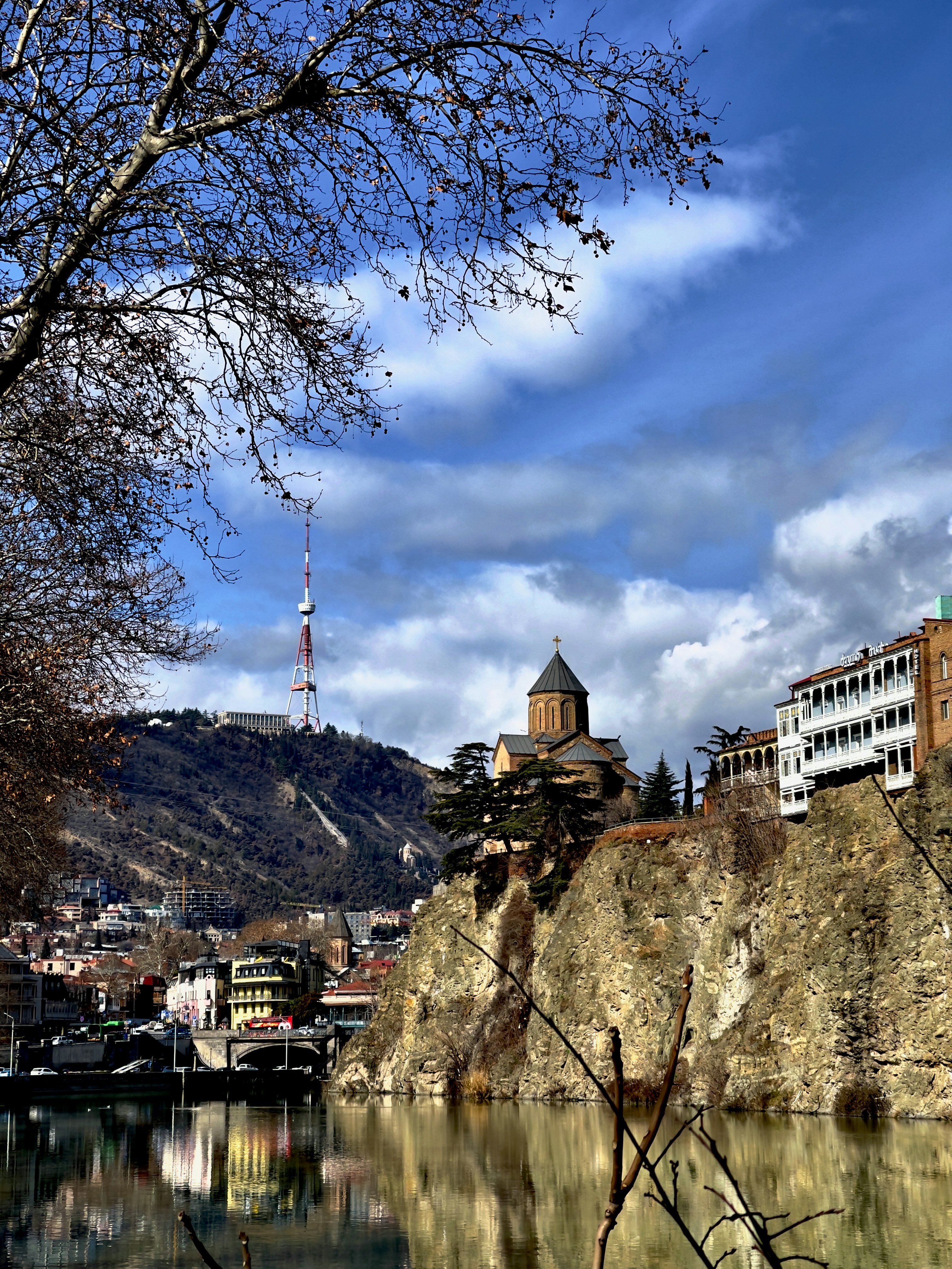 Historic church and modern tower reflect in the tranquil waters, framed by bare branches and a rugged hillside.