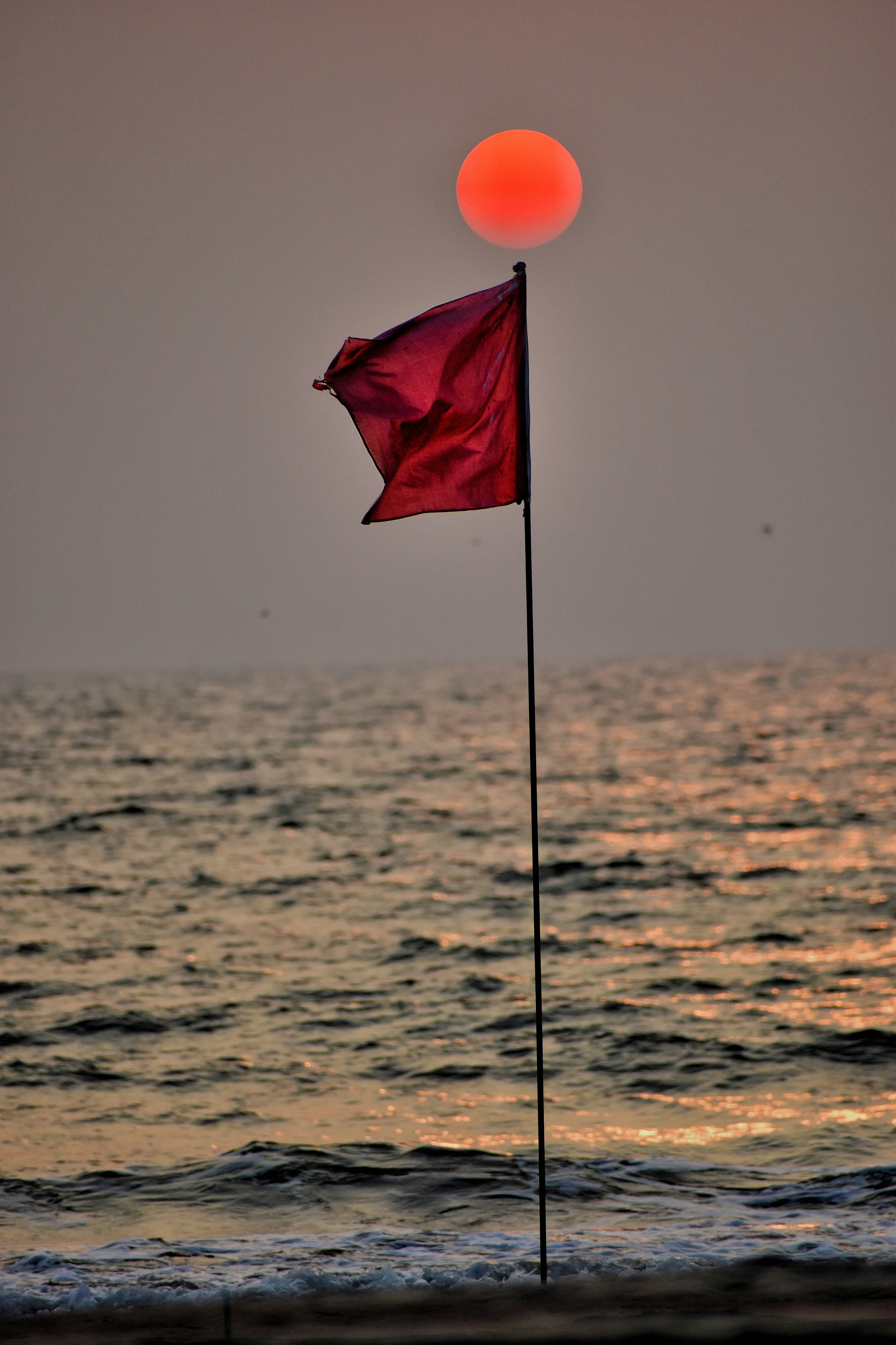 A vibrant red flag stands tall against the backdrop of a glowing sun setting over the ocean, casting a serene ambiance. The scene captures the essence of a peaceful evening by the sea.