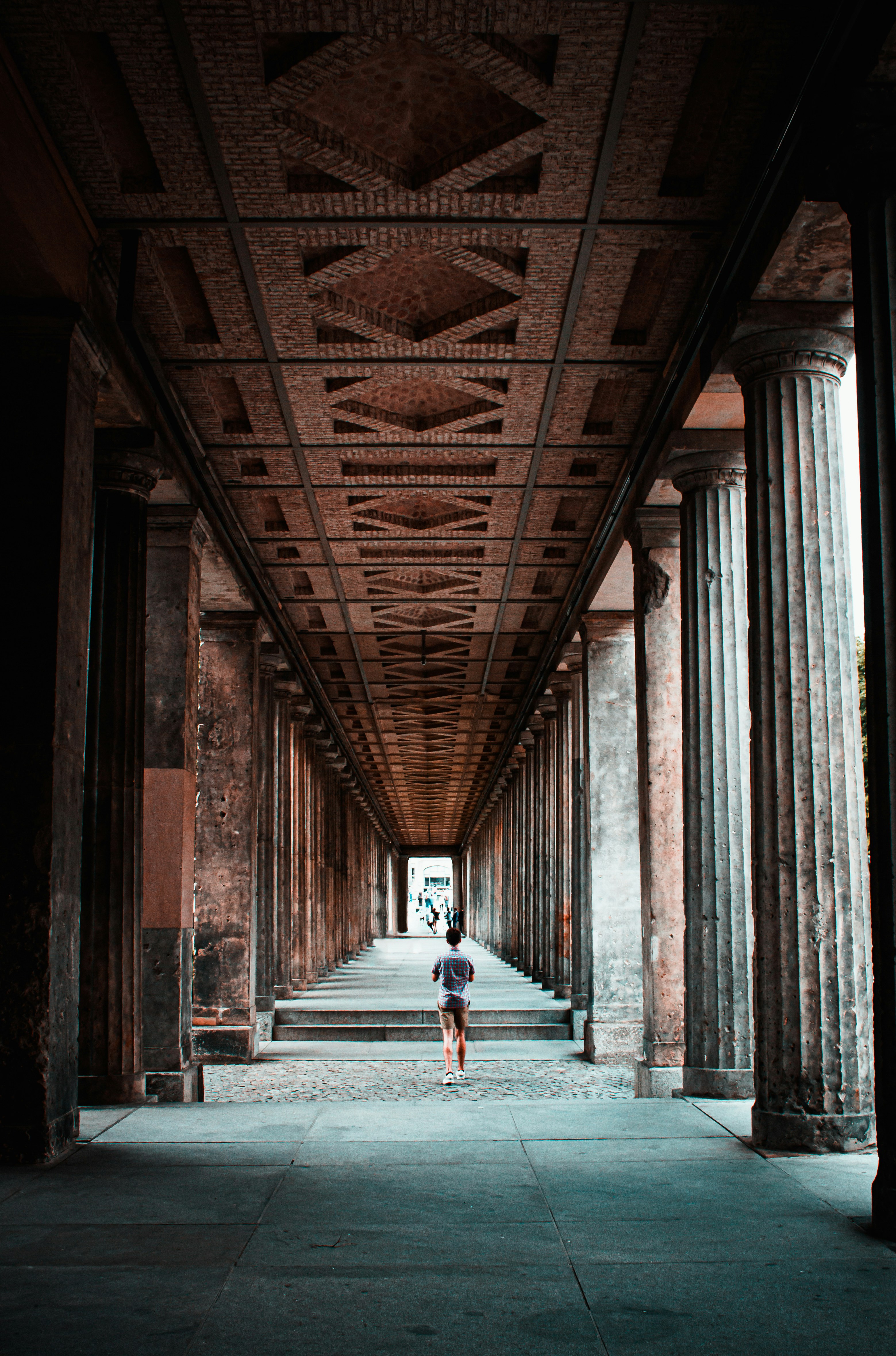 A lone figure walks through an expansive corridor lined with majestic columns and intricate ceiling patterns, leading to a bright exit in the distance.