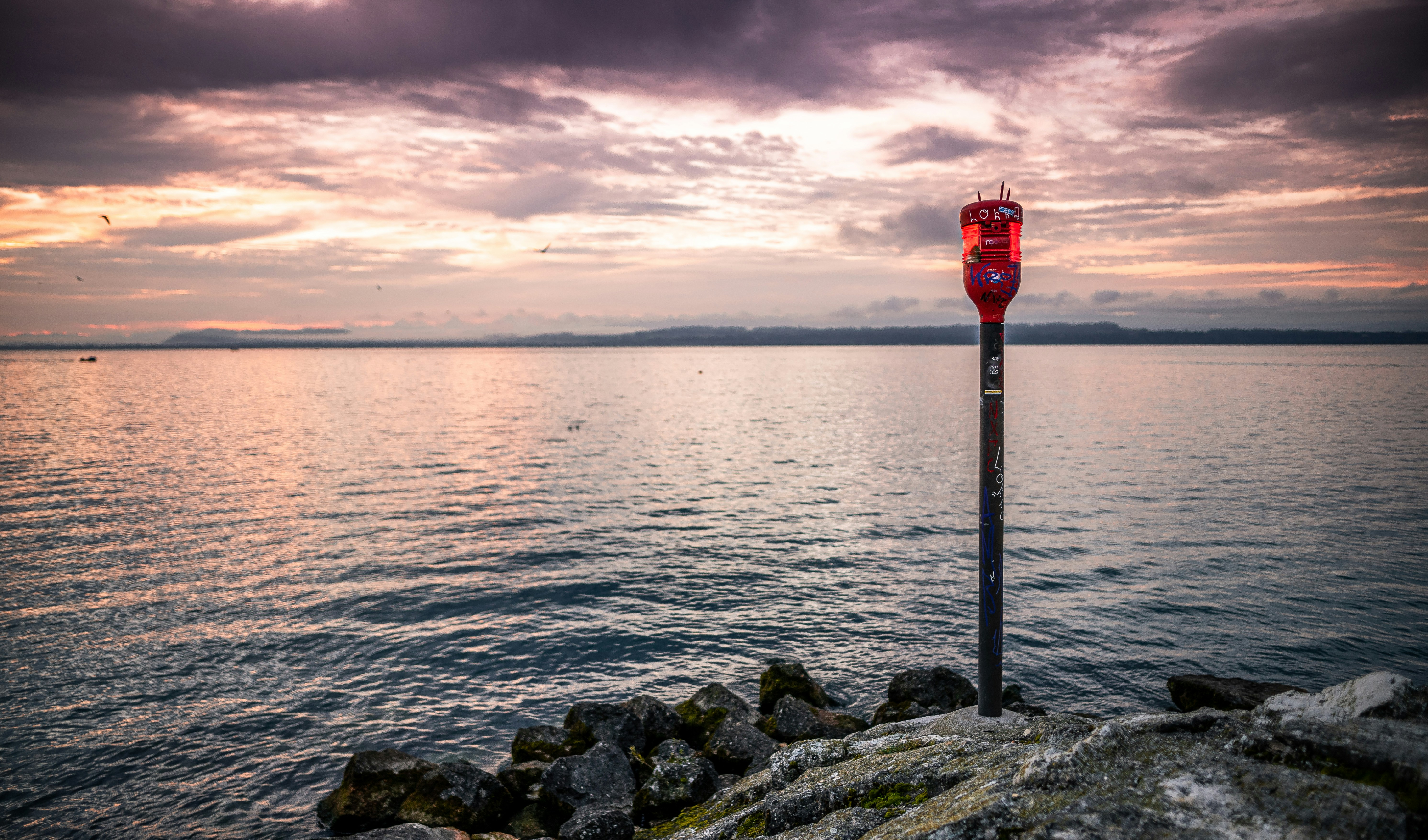 Red and white metal stand on rocky shore against a serene lake at dusk.
