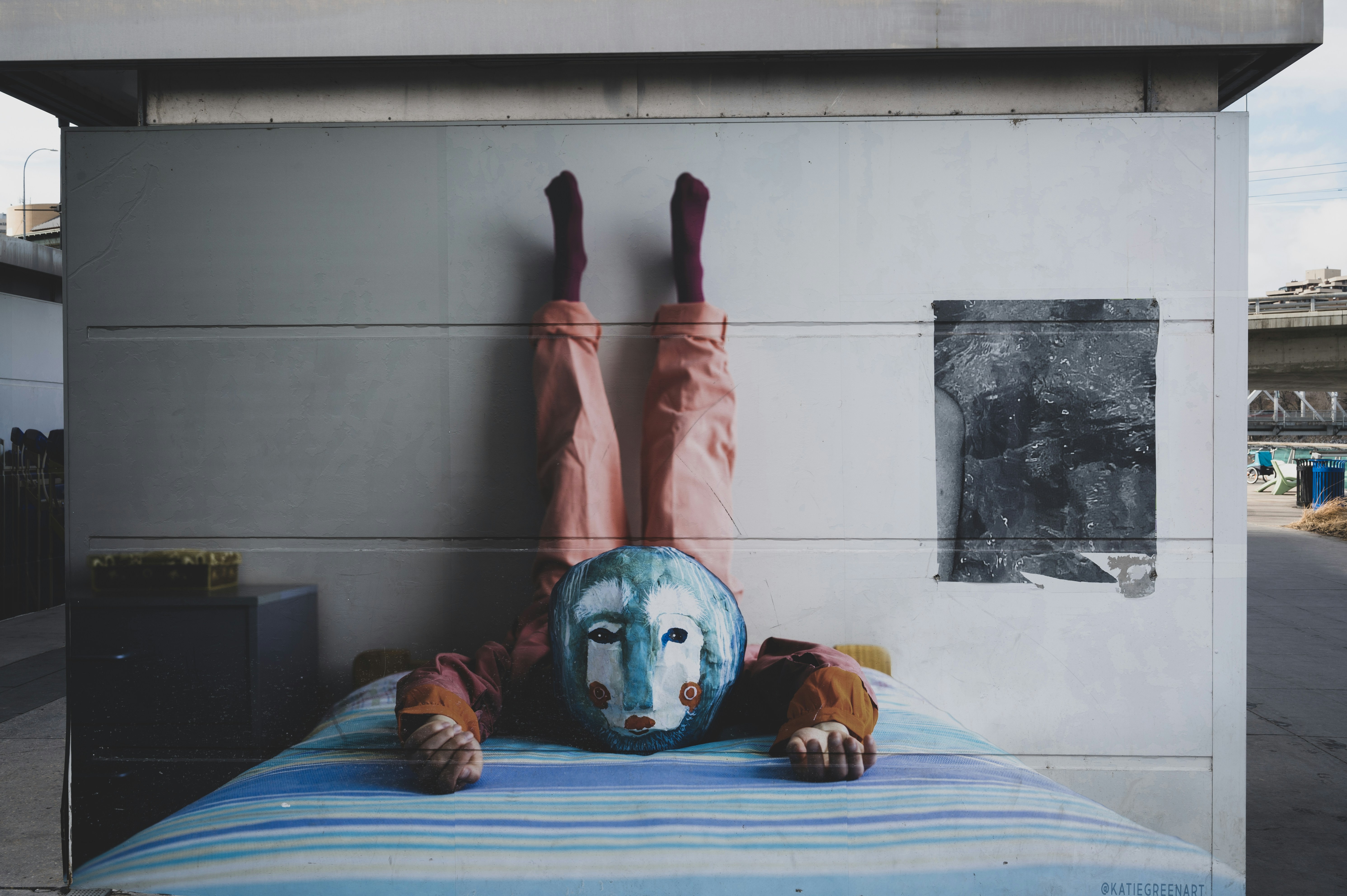 Blue-Masked Figure Upside-Down on a Striped Bed: A Surreal Street Mural