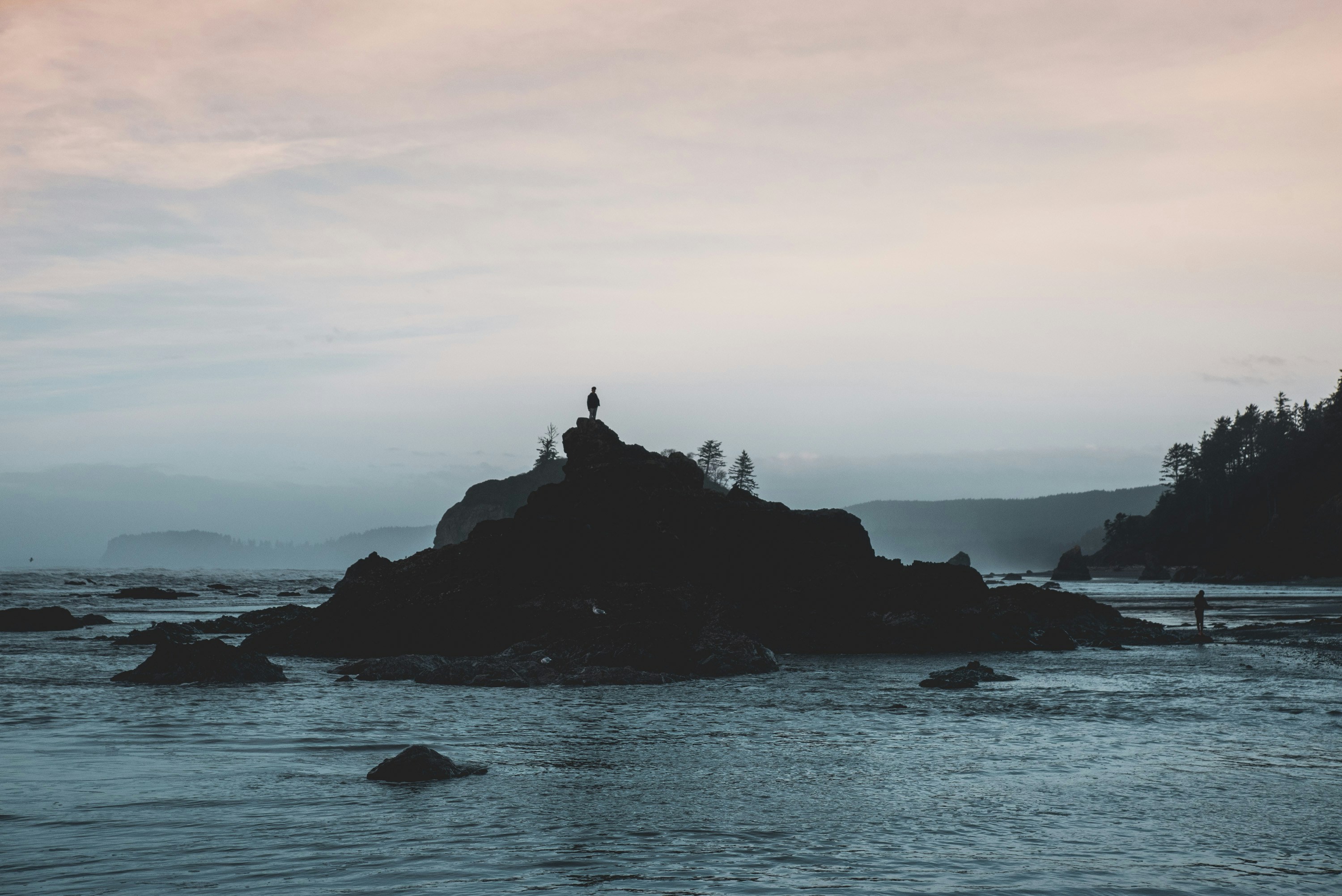 Silhouette of a rocky island against a pastel-colored sky at sunset, with calm ocean waters in the foreground.