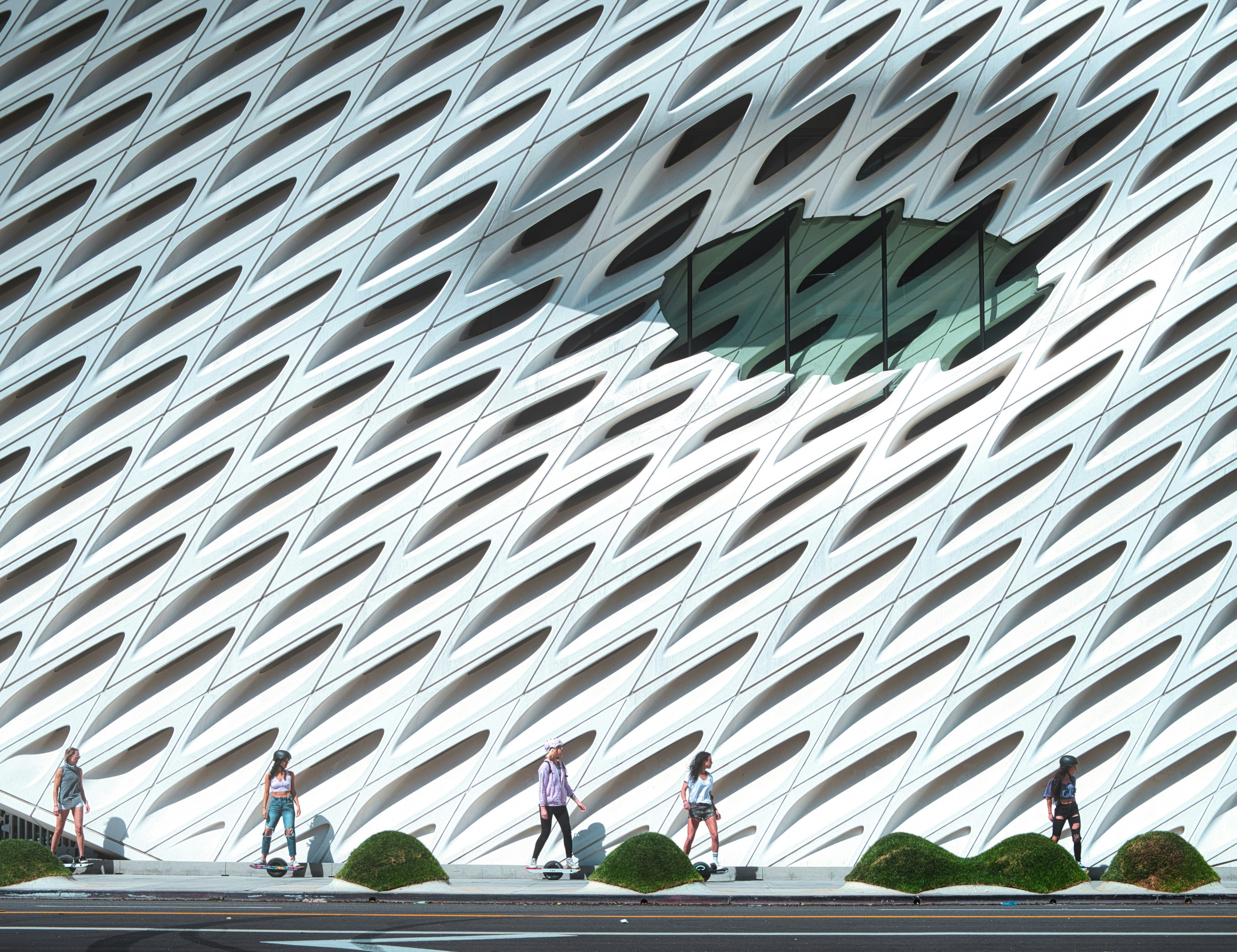 Dynamic urban scene featuring pedestrians walking along a modern building with a distinctive, cut-out window design. The facade showcases an intricate pattern that plays with light and shadow.