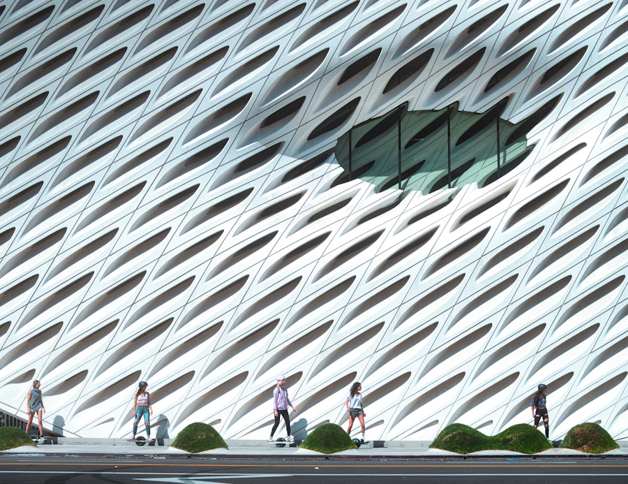 Five skateboarders lined up along the base of a sweeping white lattice facade, with a dark green window band breaking the pattern above. Strong shadows and repeating diamond-shaped openings create a graphic urban scene.