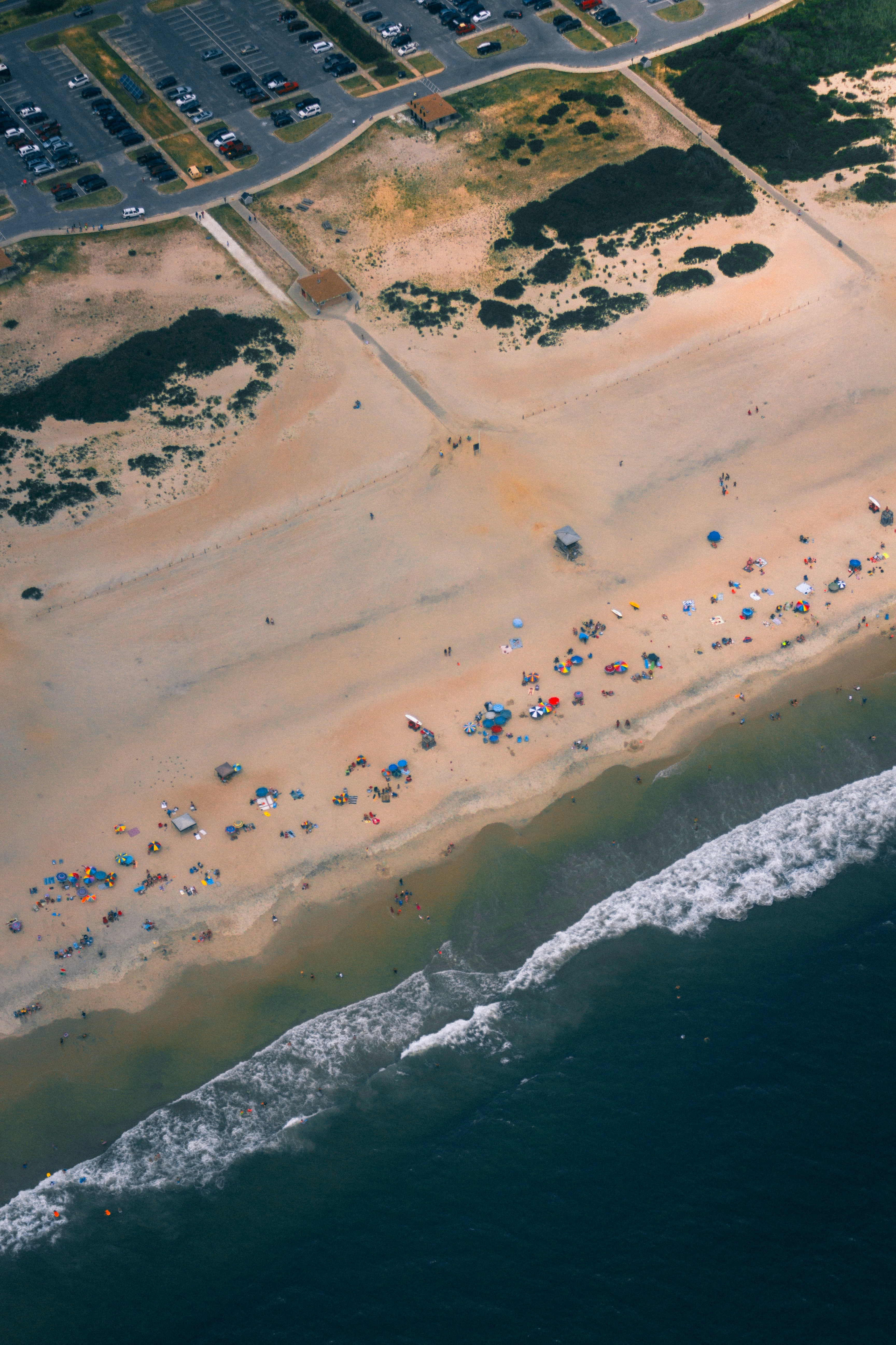 Aerial view of a sandy beach bustling with colorful umbrellas and sunbathers, bordered by gentle waves and lush greenery.