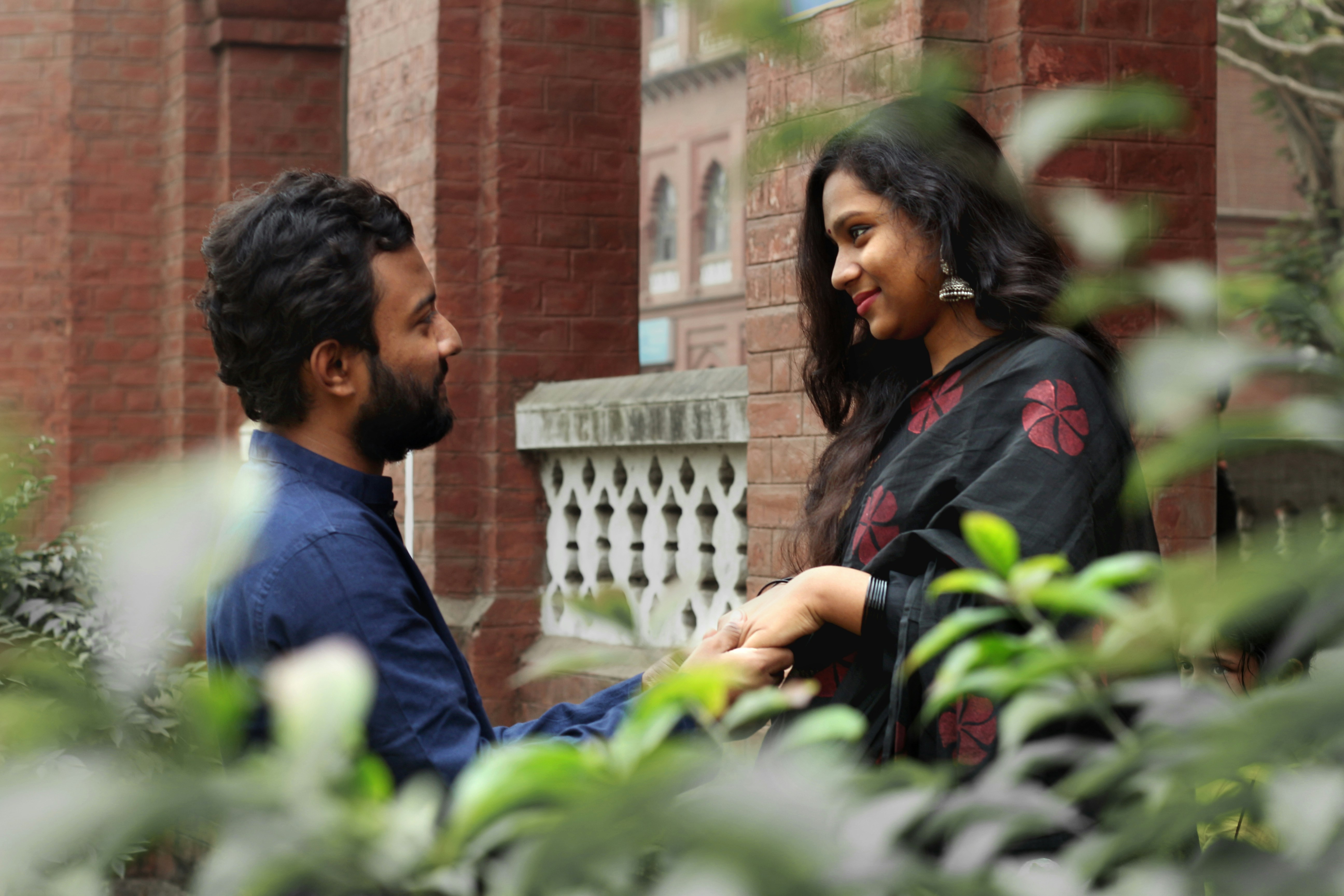 Man and woman sharing a moment outdoors, framed by lush foliage against a brick backdrop.