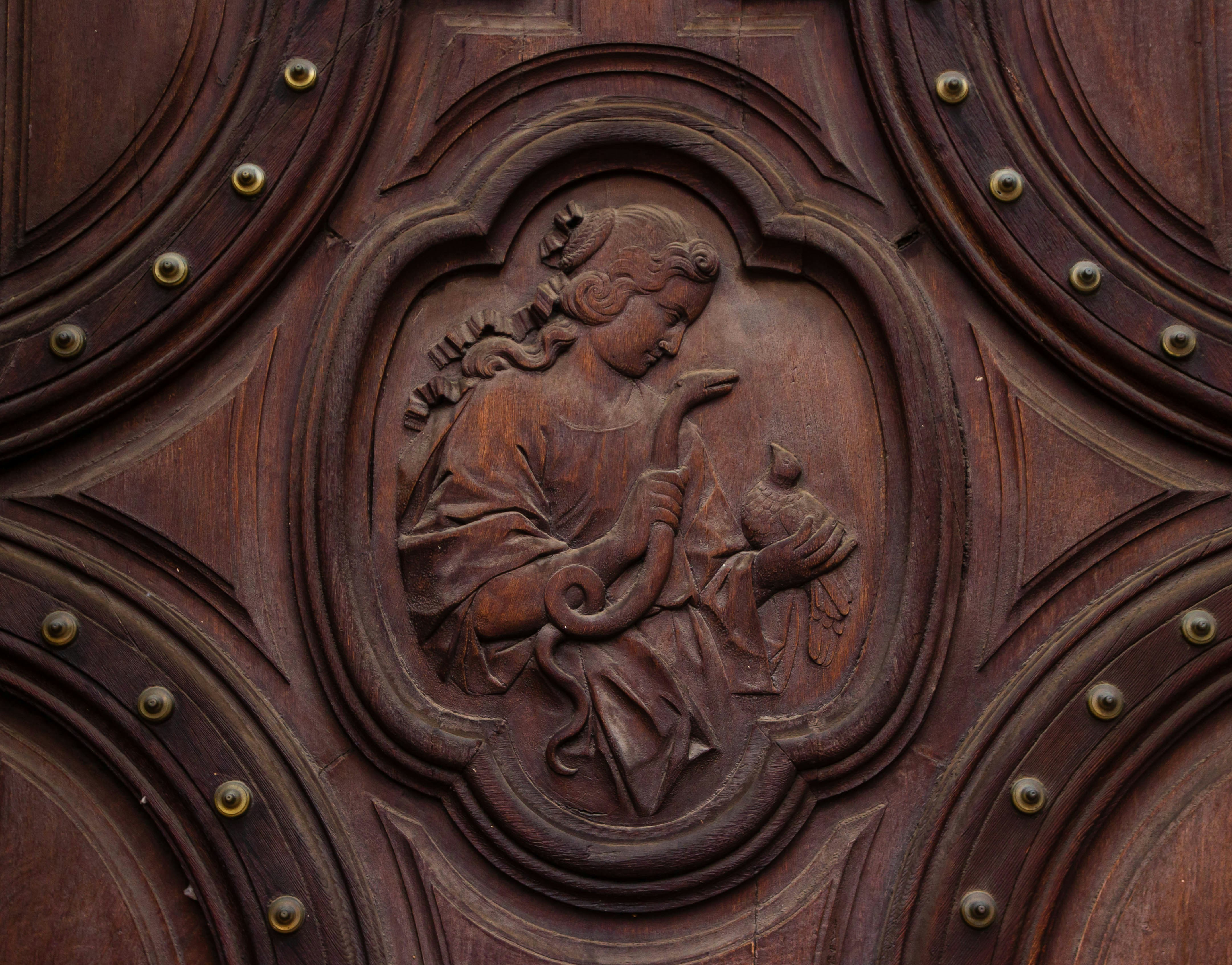 Intricate wooden relief depicting a figure holding a snake and a bird, surrounded by ornate framing. The craftsmanship highlights the artistry of woodwork.