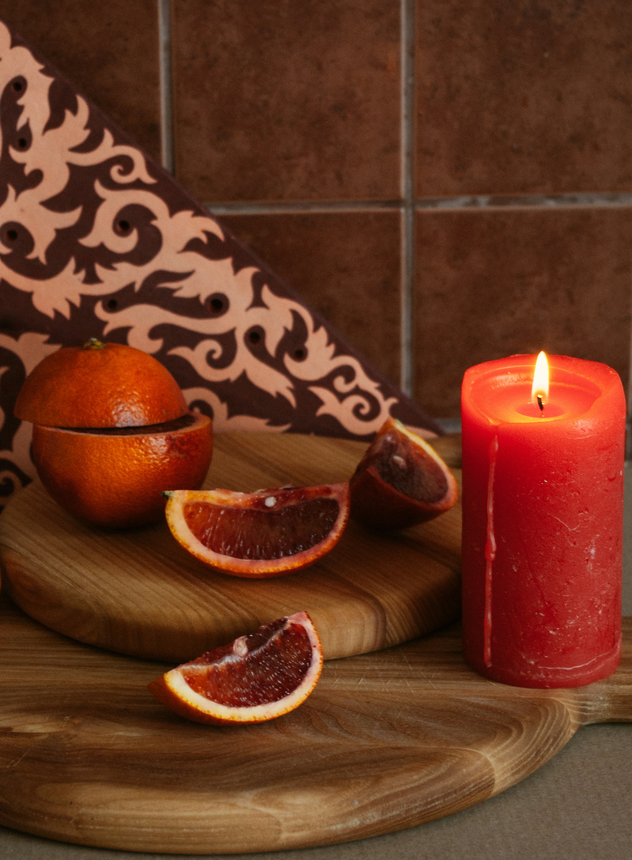 Blood oranges and a red candle arranged on wooden surfaces, set against a patterned backdrop. The scene exudes warmth and texture.