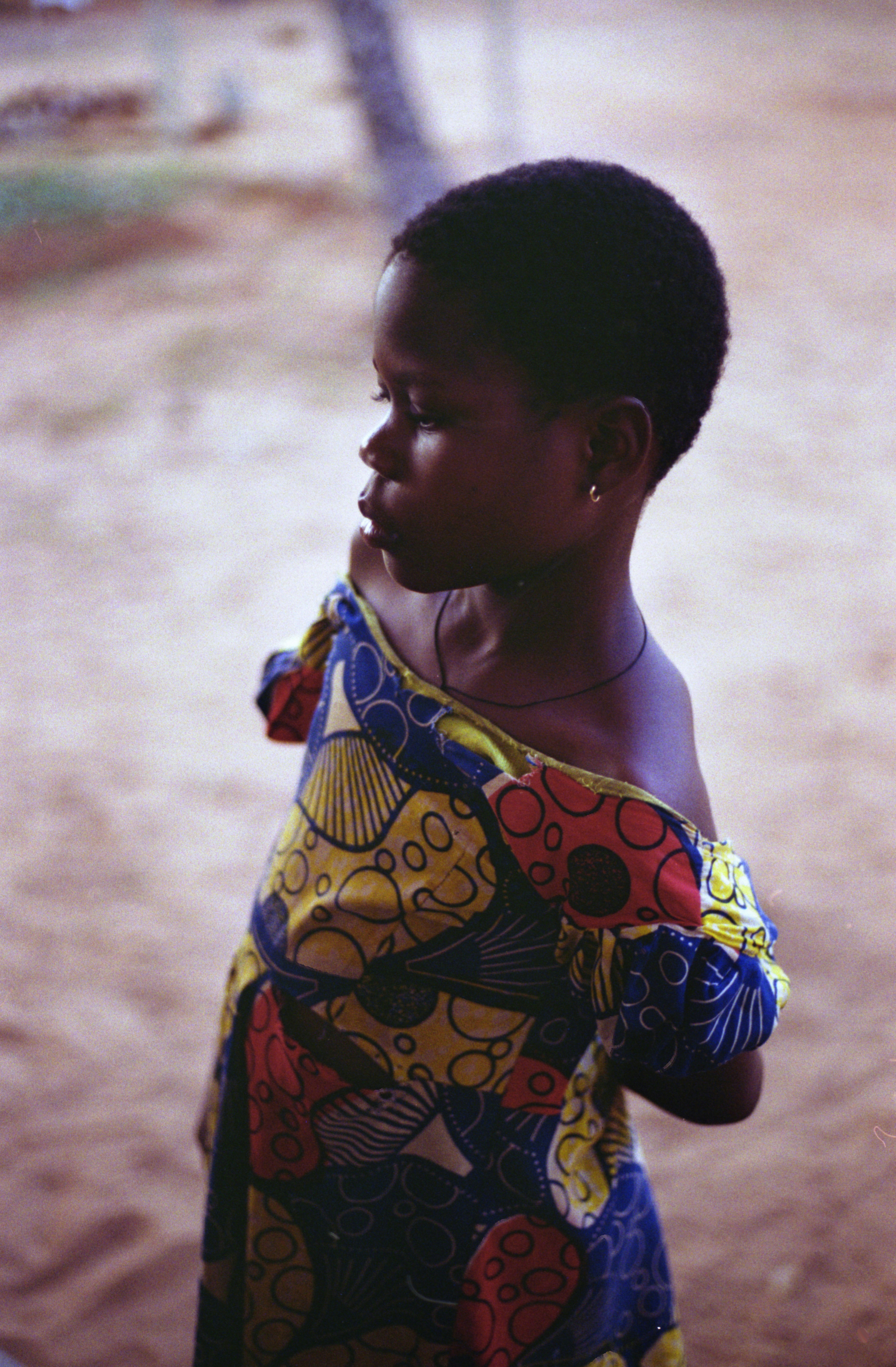 A young girl in a colorful, patterned dress stands thoughtfully, her profile illuminated by soft light. The background hints at a natural setting, enhancing the moment's tranquility.