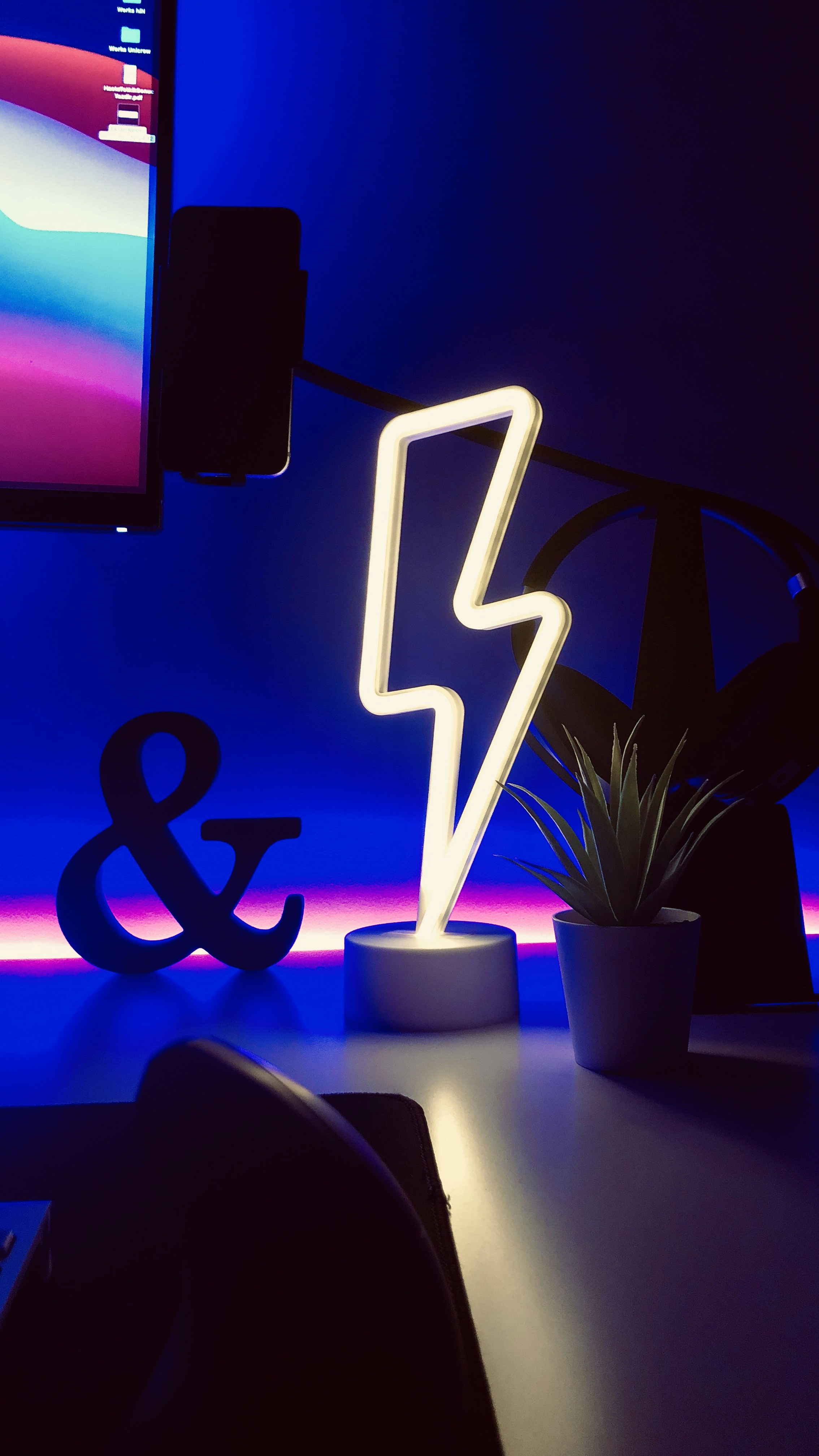 A neon lightning bolt lamp stands prominently on a desk, accompanied by a decorative plant and an ampersand symbol, all set against a vibrant blue backdrop.