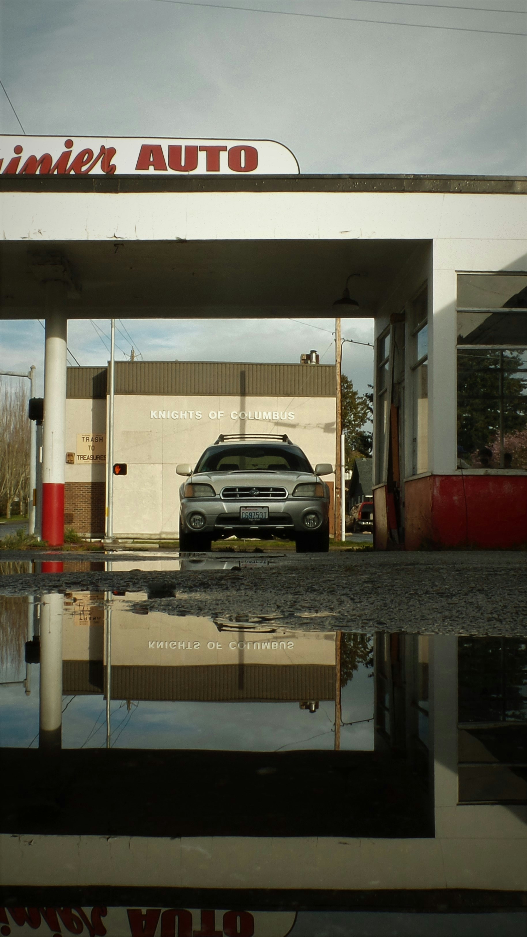 Silver SUV under a shelter with a reflected car and building sign, framed by a roadside auto service; a calm, symmetrical composition with puddle reflection.