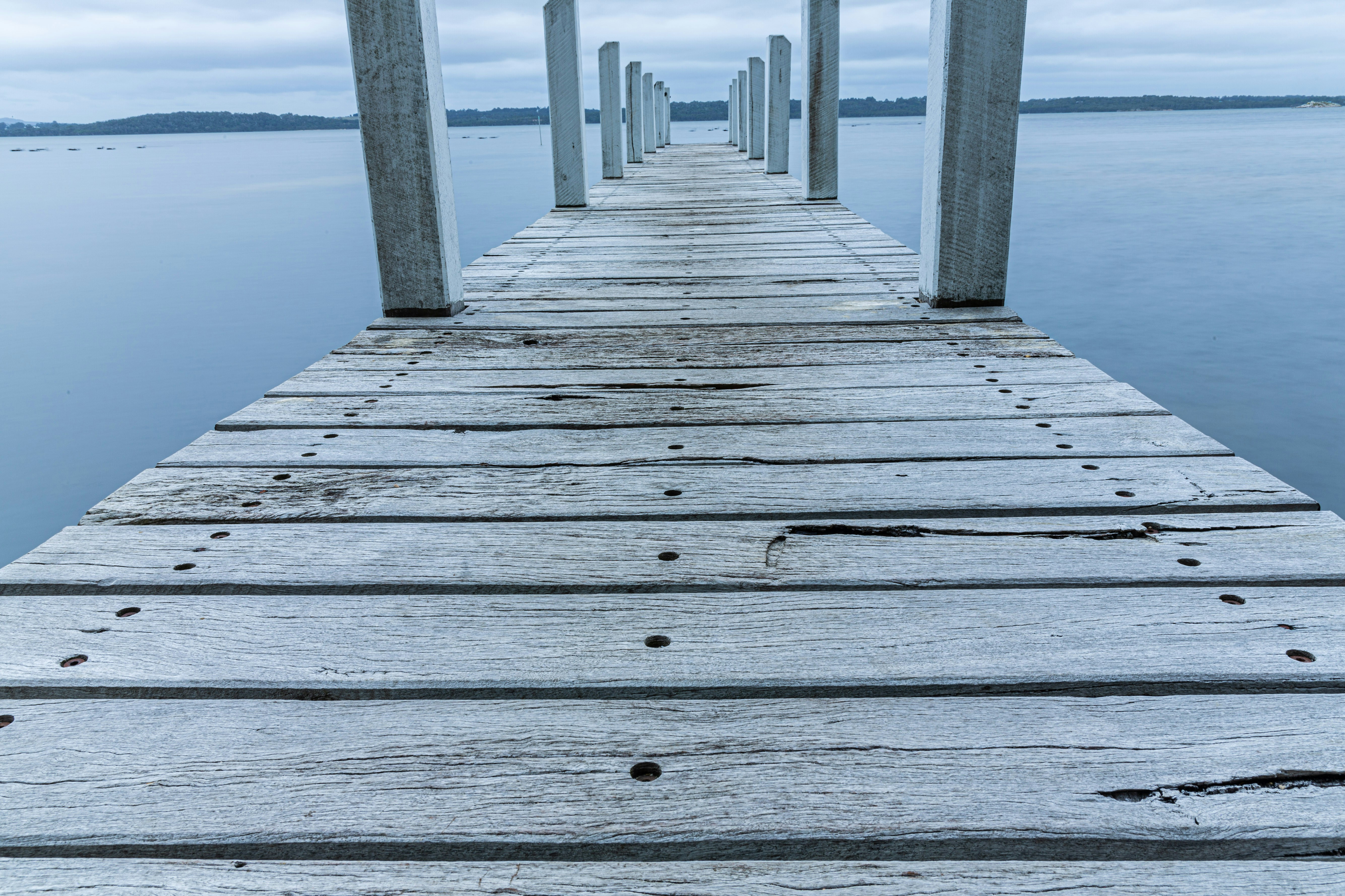 Weathered wooden pier extending into calm waters under a soft gray sky. The scene evokes a sense of tranquility and reflection.