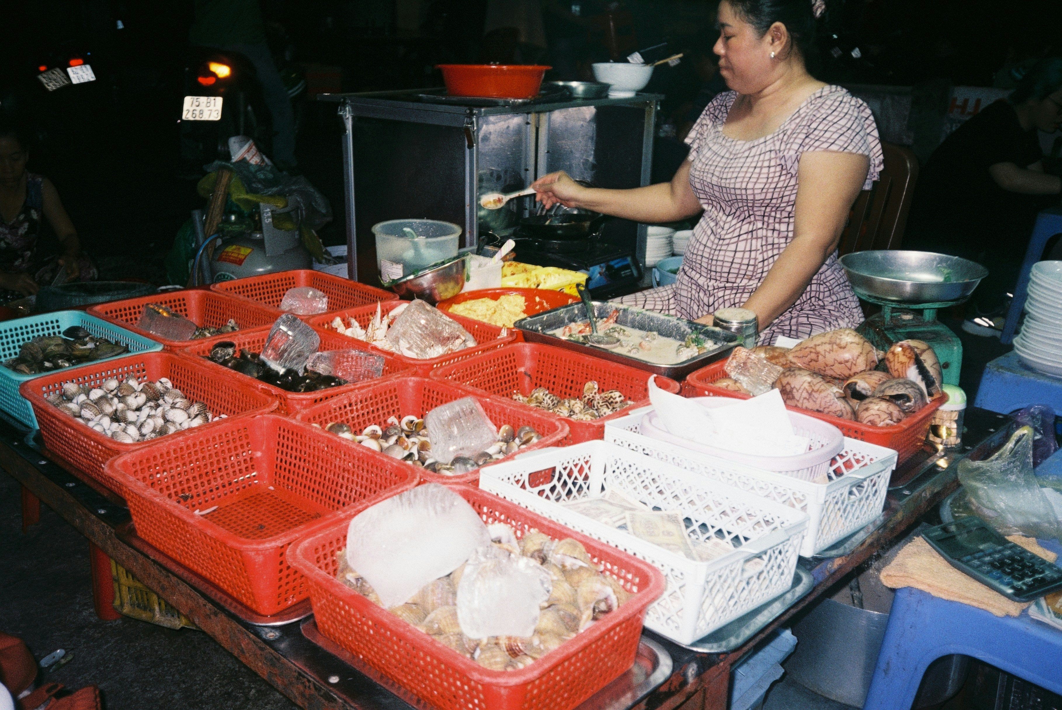 A bustling street vendor prepares a variety of seafood dishes, showcasing colorful baskets filled with fresh ingredients. The scene captures the vibrant atmosphere of a night market.
