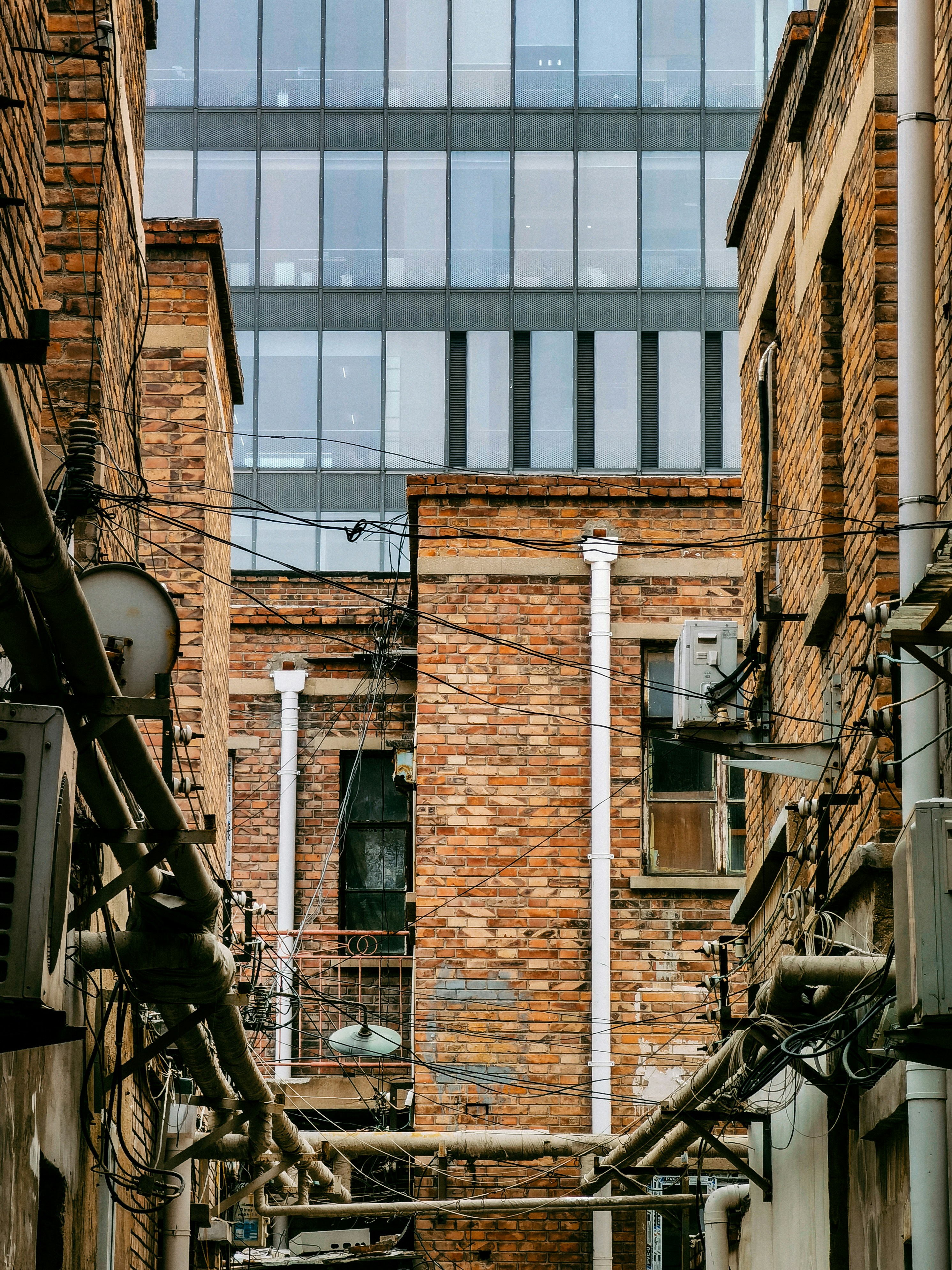 Narrow alleyway flanked by brick buildings and overhead pipes, contrasting with a sleek glass facade above. 