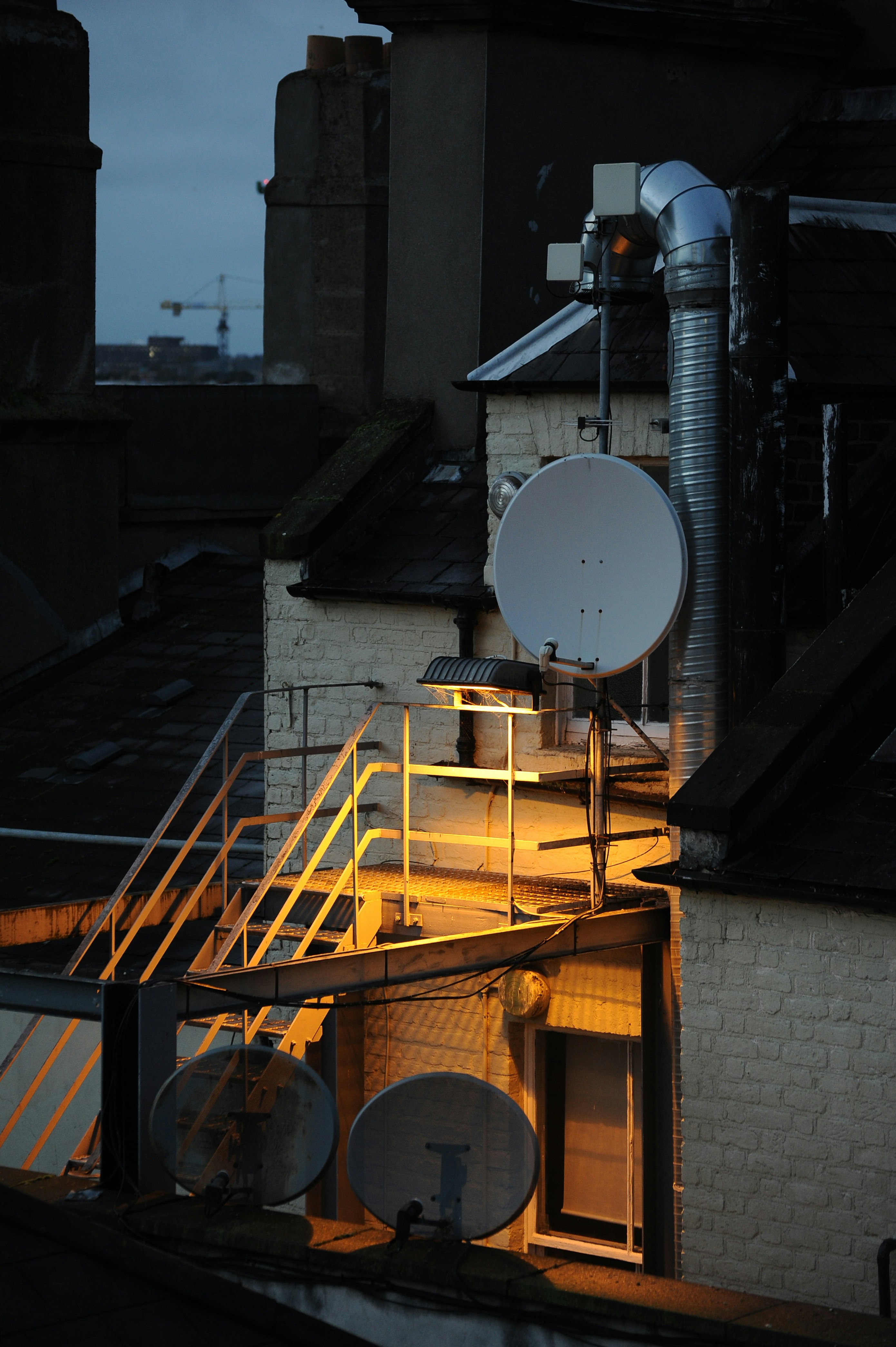 A rooftop scene featuring satellite dishes and a metallic staircase illuminated by warm light, set against a twilight sky.