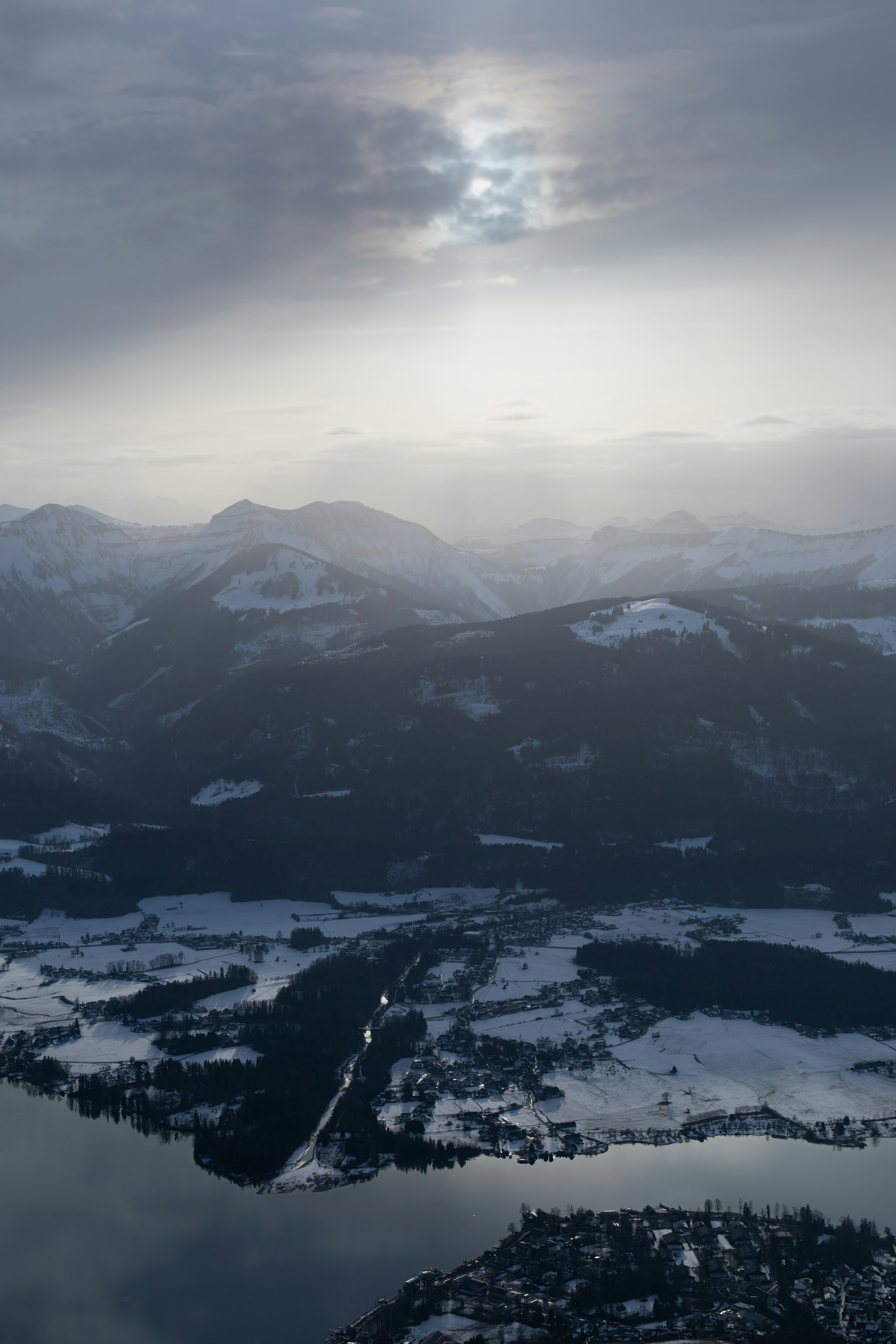 Snow-covered mountains loom over a tranquil lake and village, shrouded in soft winter light. The scene evokes a sense of peace and solitude.