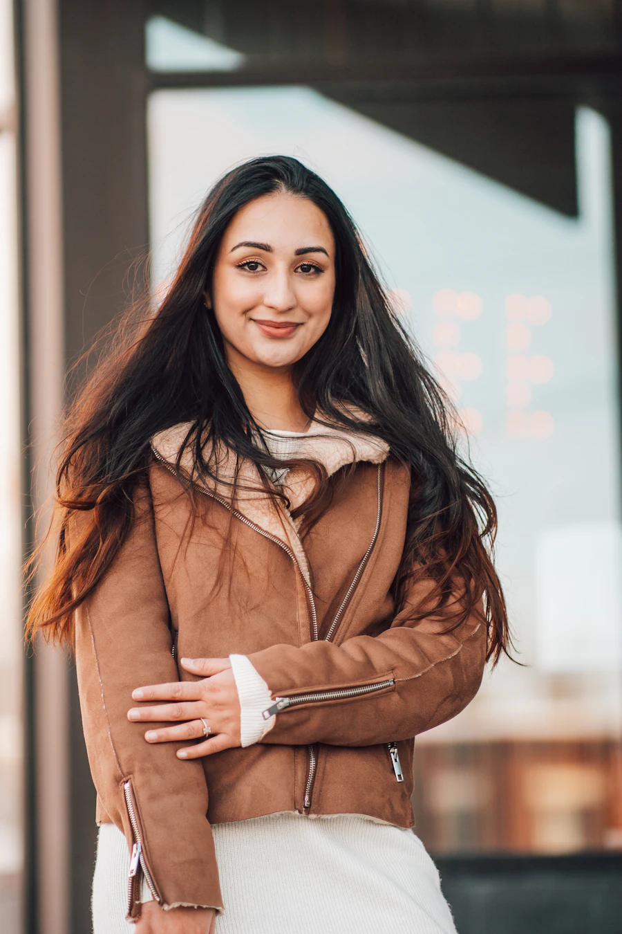 Young woman with long dark hair standing outdoors in a brown jacket and cream sweater, smiling at the camera. Soft blurred background with warm evening light.