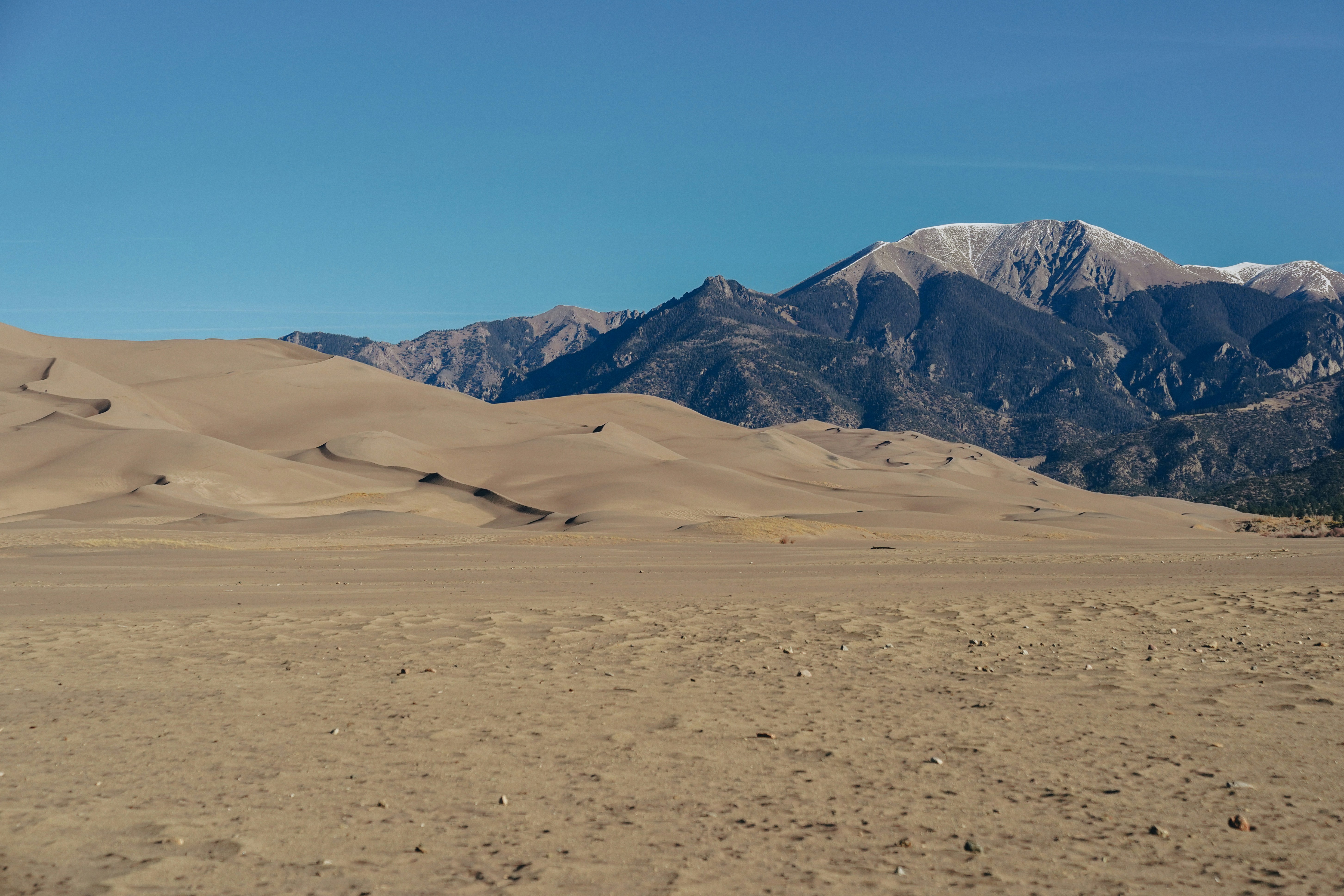 Vast sandy dunes stretch across the foreground, leading to a majestic mountain range under a clear blue sky.