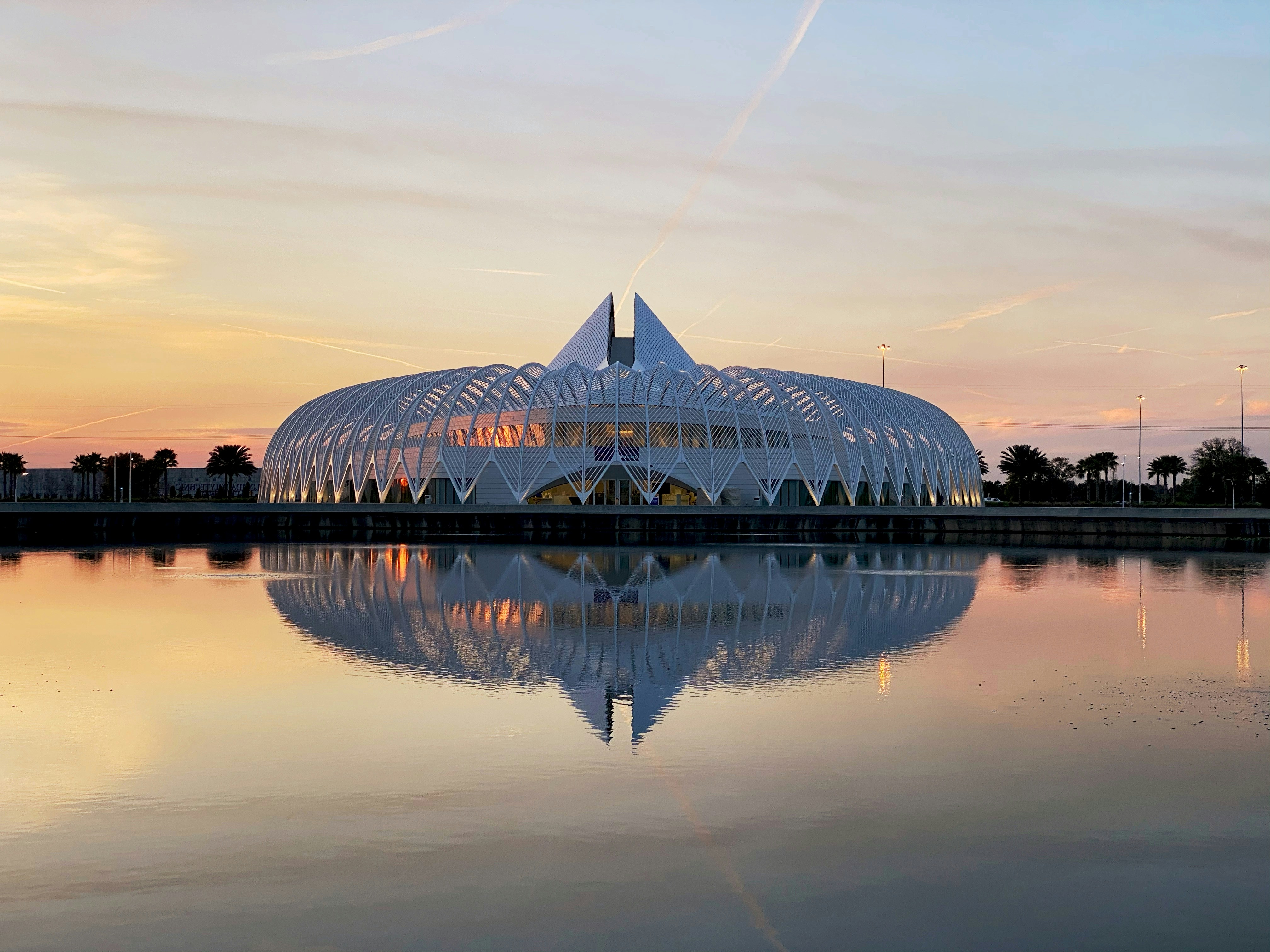 Modern white building with a geometric design reflected in a calm body of water at sunset.