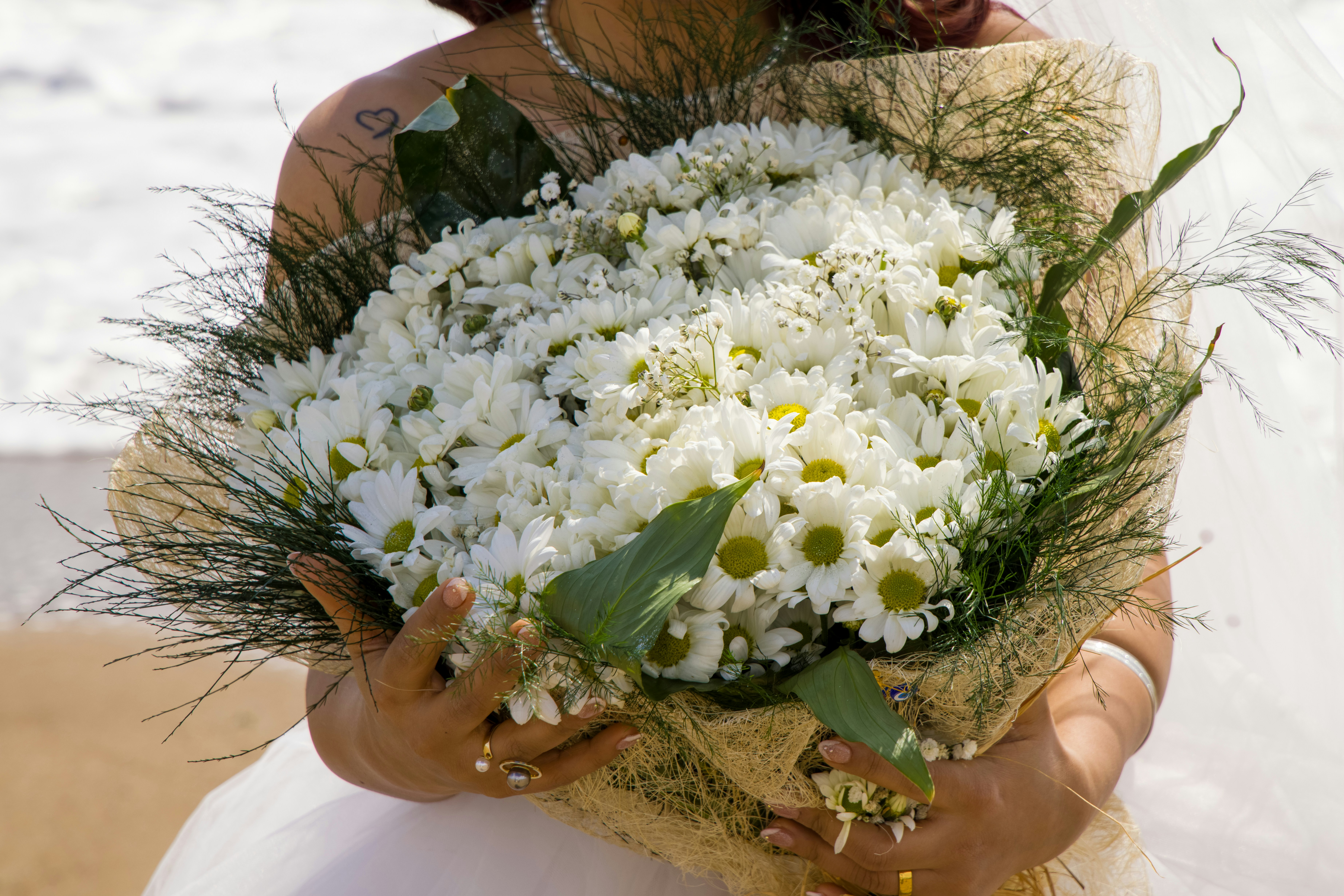A bride holds a lush bouquet of white daisies and greenery, radiating joy and purity against a soft, blurred background of the beach.