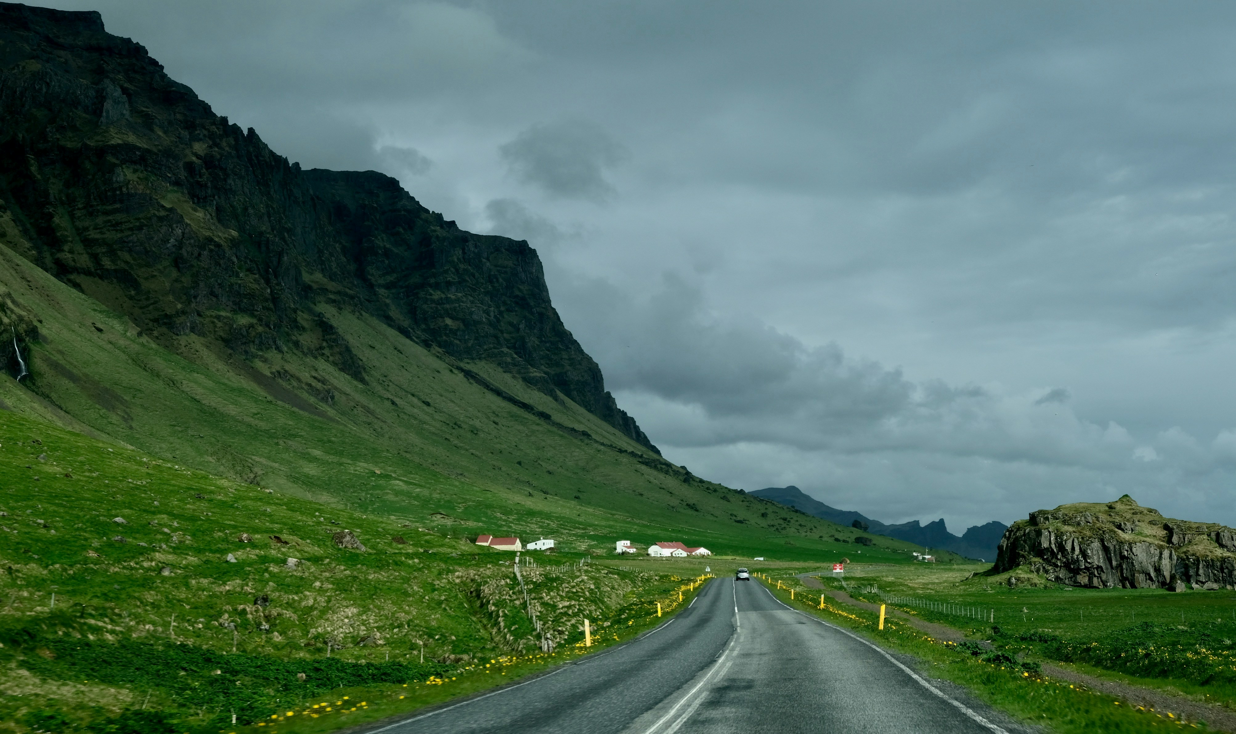 Winding road leads through lush green hills beneath a dramatic sky, with distant mountains framing the scene. Small farmhouses dot the landscape.