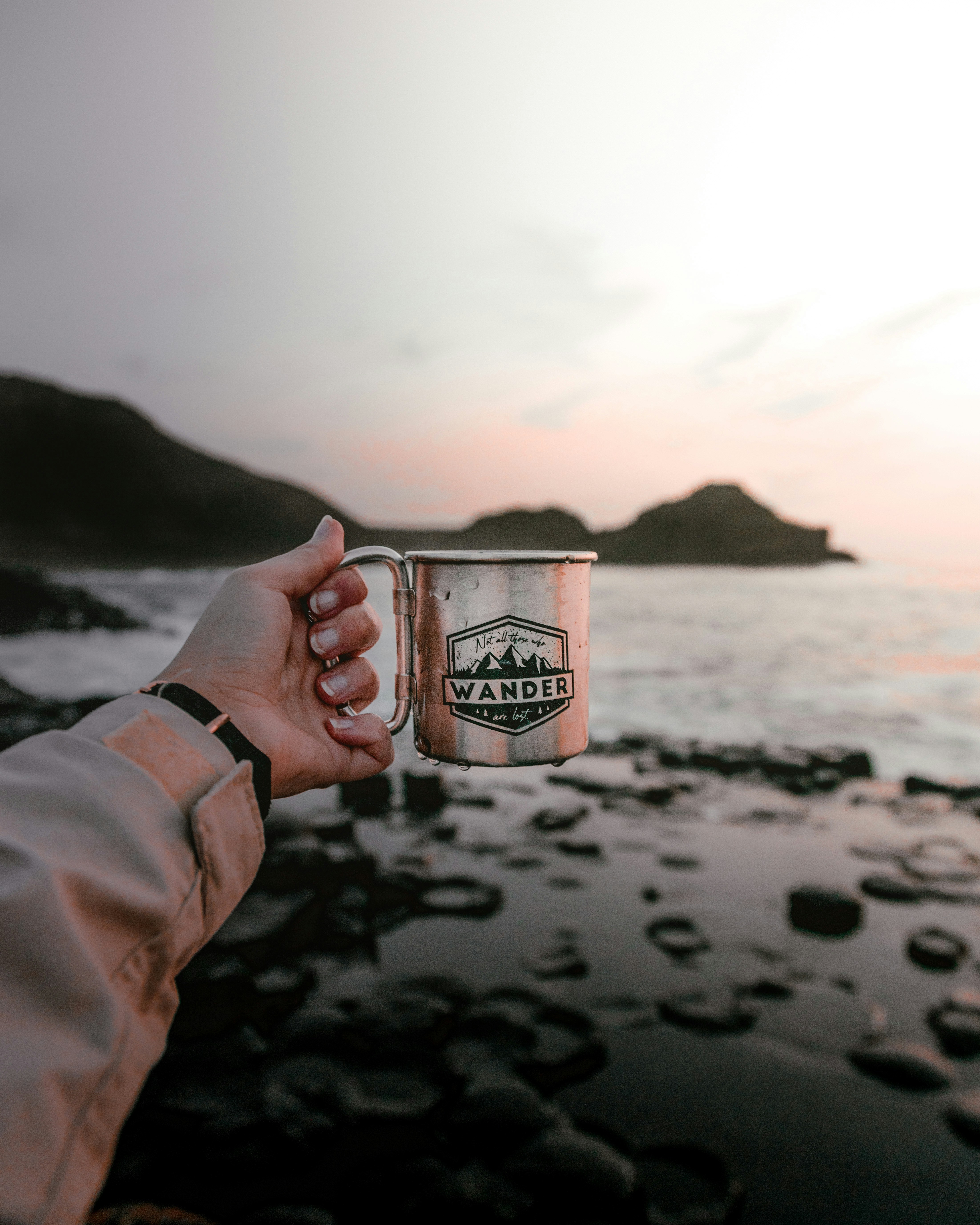Hand holding a stainless steel mug with 'WANDER' logo against a serene coastal backdrop during sunset.