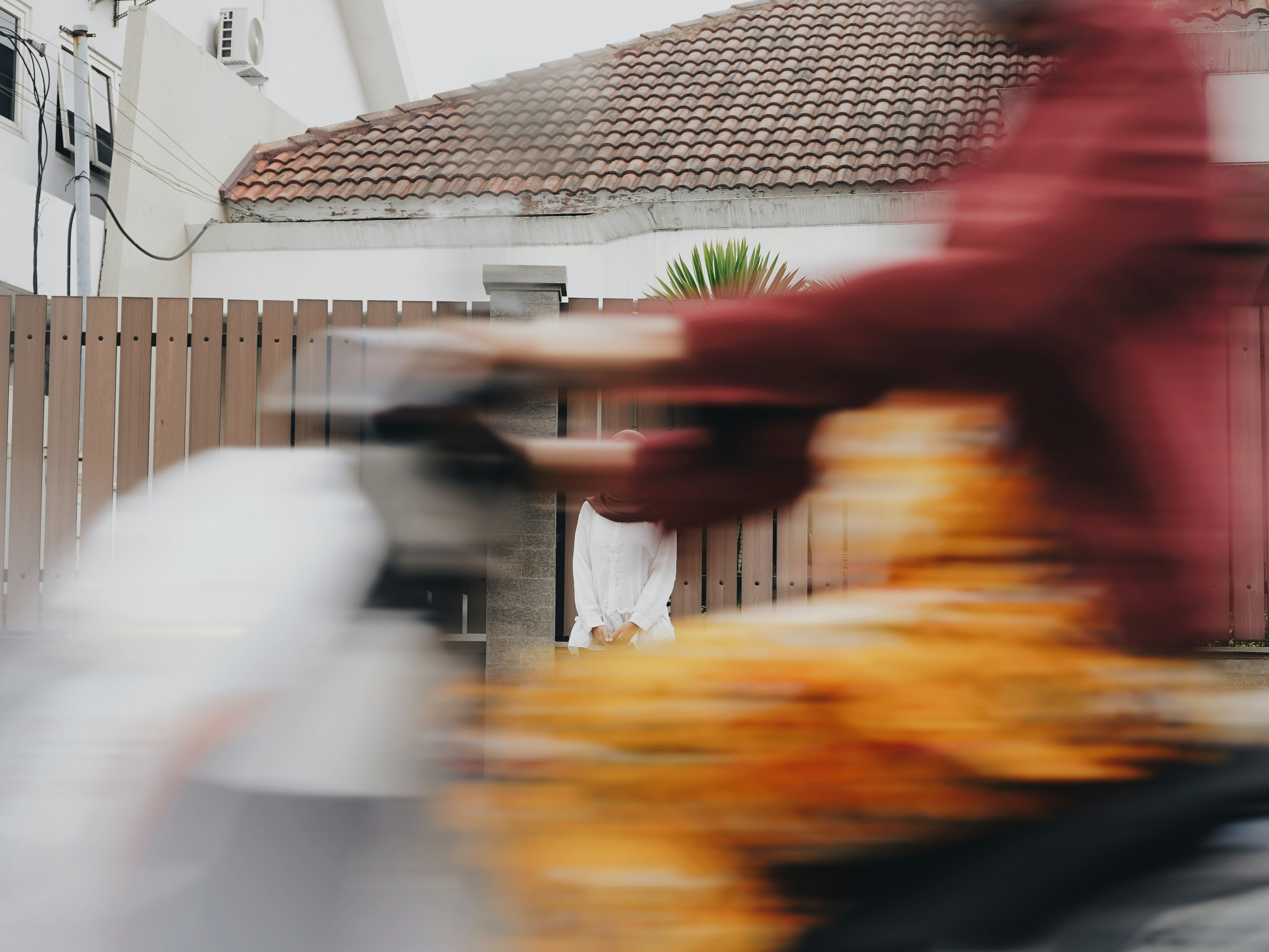 Blurred figure on a scooter passing by a rustic fence and tiled roof.