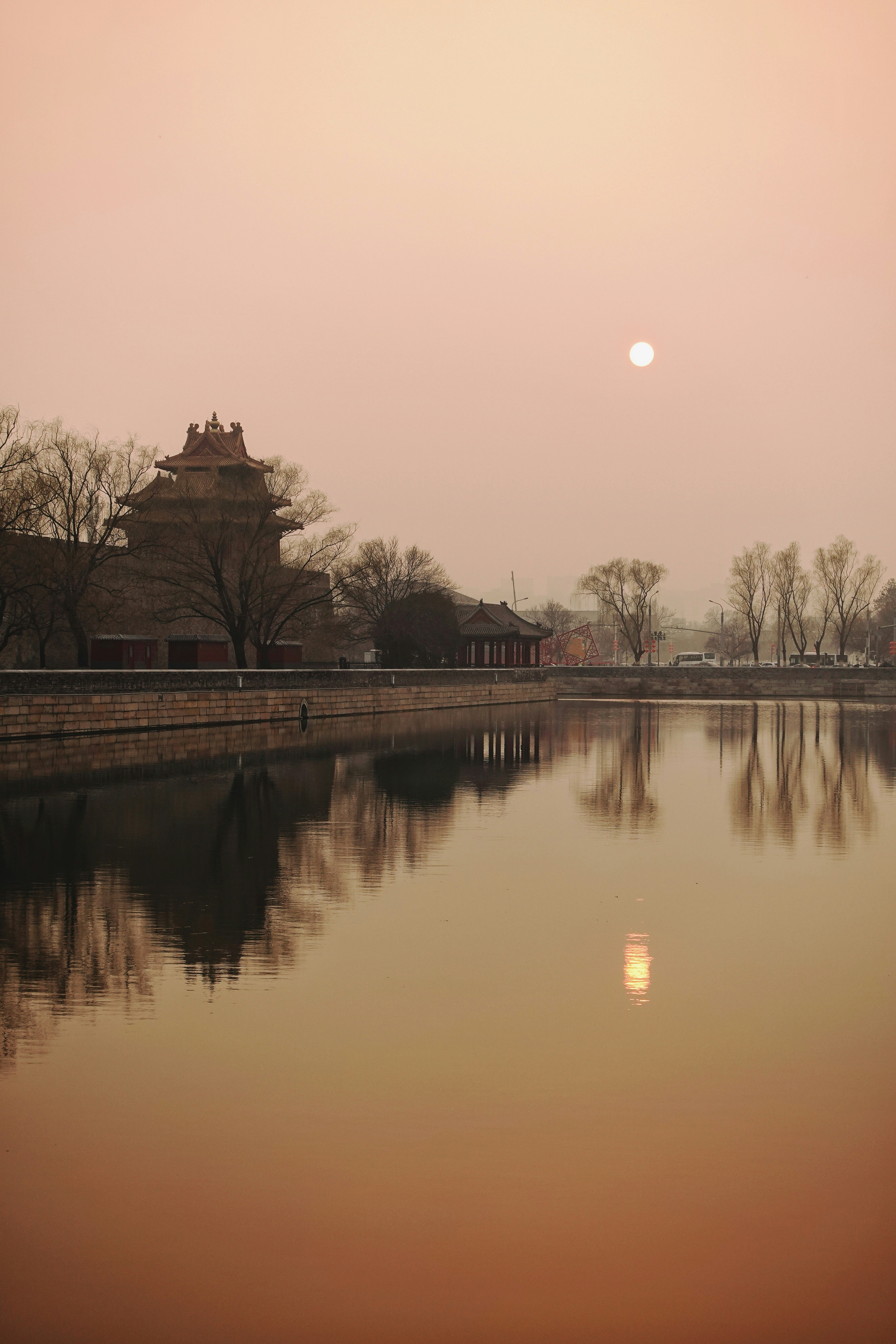 Serene view of a historical structure reflecting in calm waters at sunset, enveloped in a soft haze.