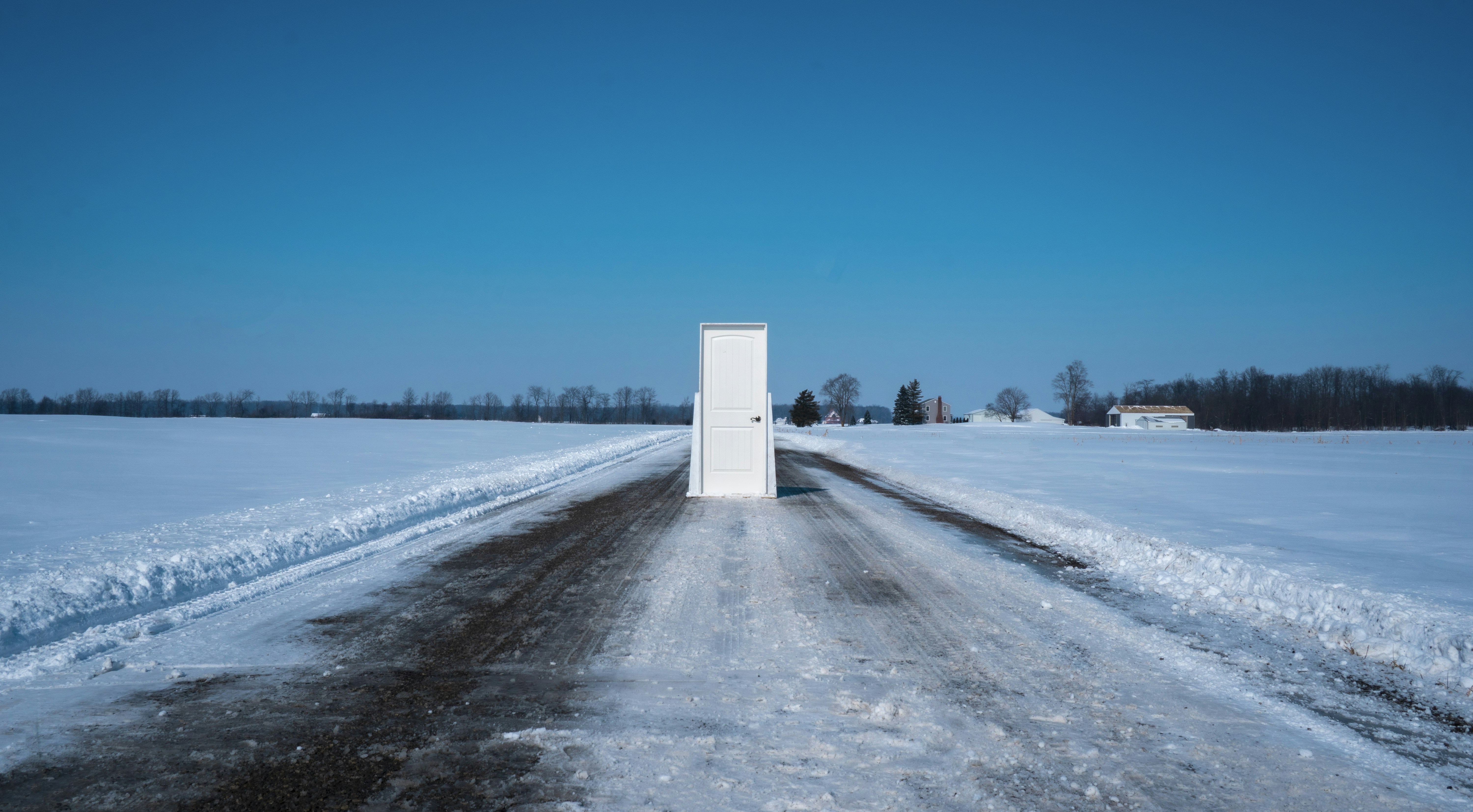A solitary white door stands in the middle of a snow-covered road, surrounded by vast fields under a clear blue sky.