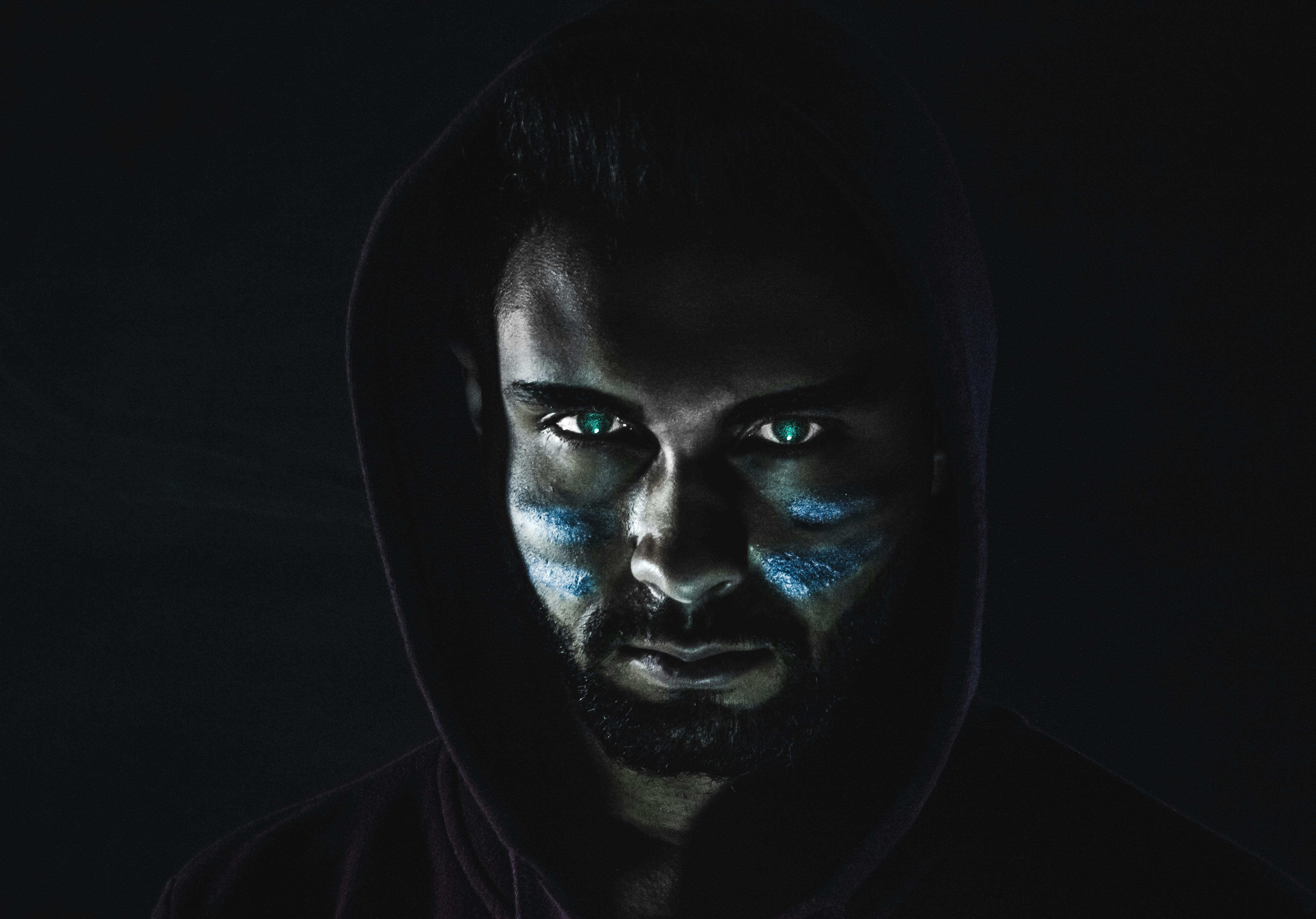 Portrait of a person in a hood with face paint, eyes illuminated in low light.