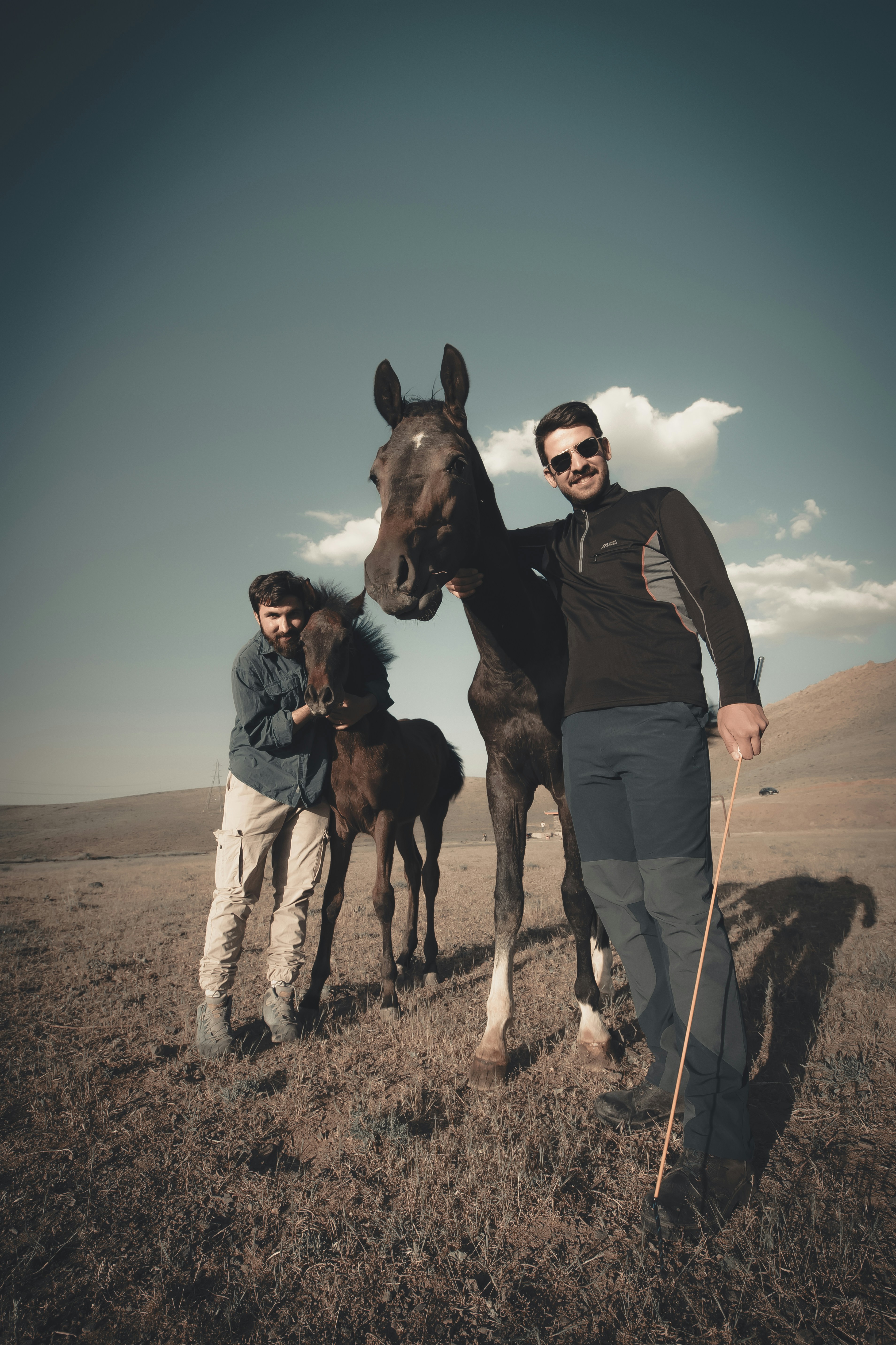 Two men interacting with a horse and its foal in a vast grassland under a clear sky.